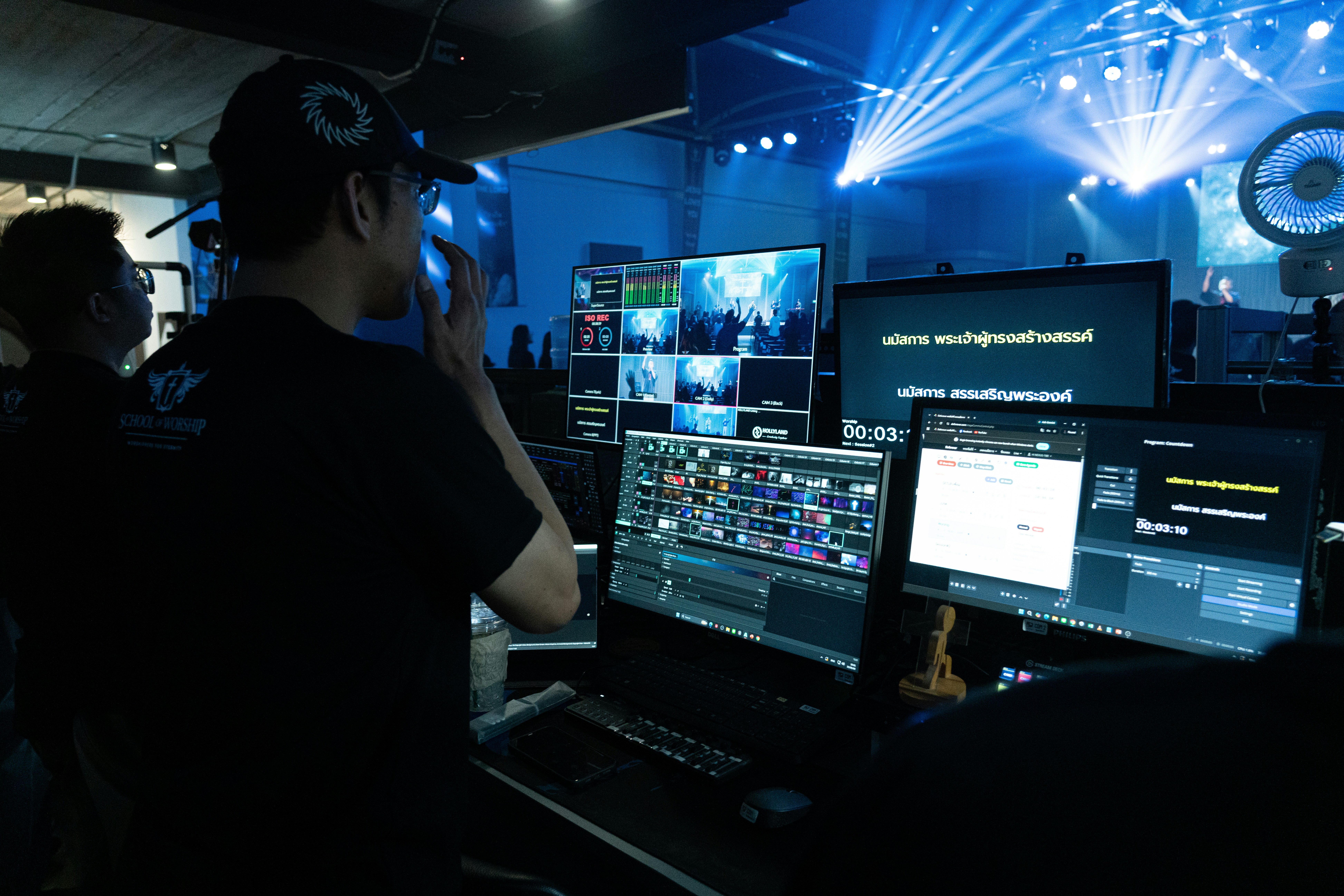 A live control room with multiple monitor walls displaying feeds and timelines, illuminated by blue stage lighting. This is a behind-the-scenes view of a production setup, likely a broadcast or event control area.