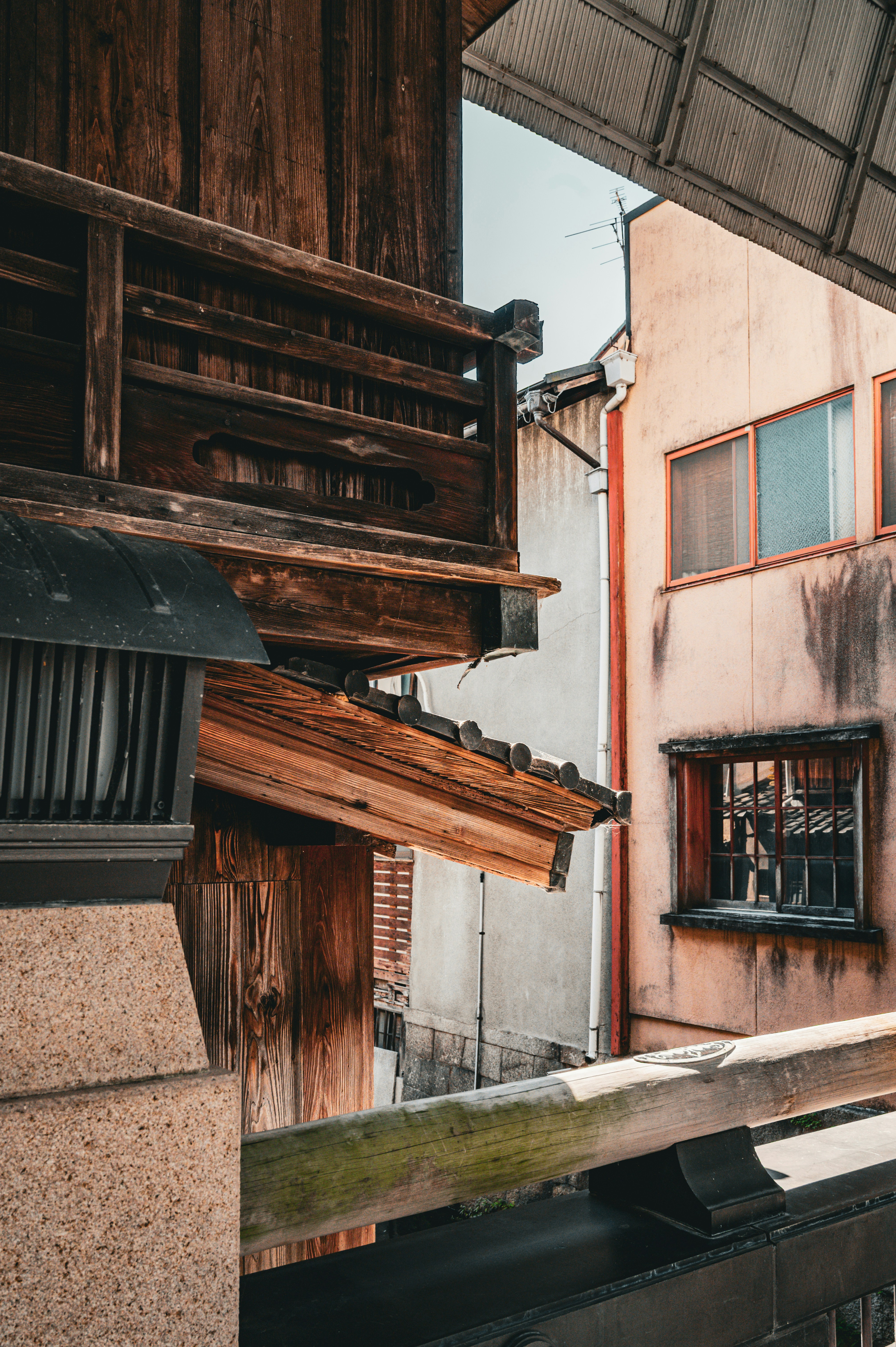 Close-up of a weathered wooden balcony and adjacent building walls in an urban alley, revealing traditional architecture and textures. A hint of modern window frames and mossy ledge adds contrast.