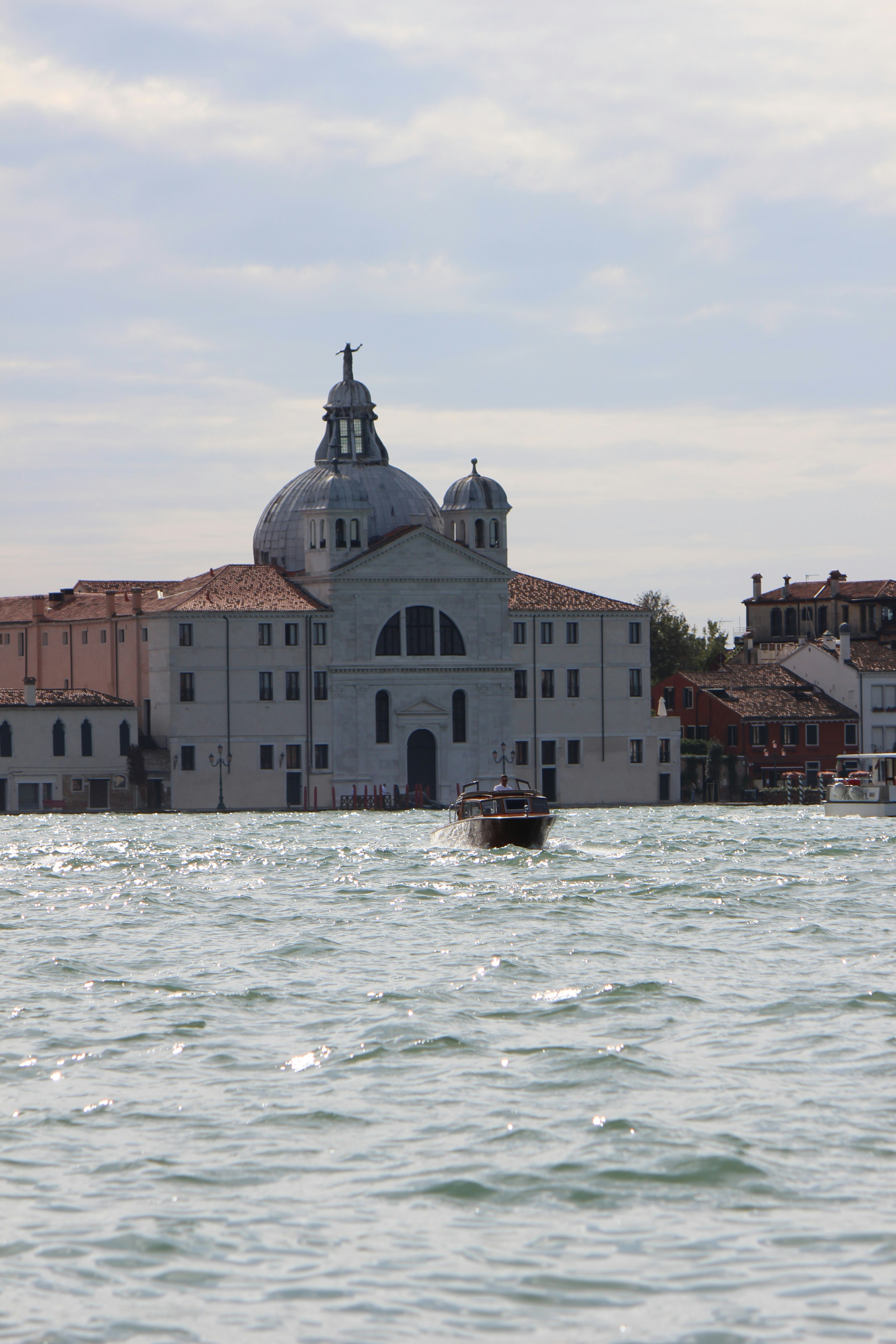 White waterfront churchfront with domed towers sits across a shimmering canal, a small boat gliding toward its steps amid sunlit waves.