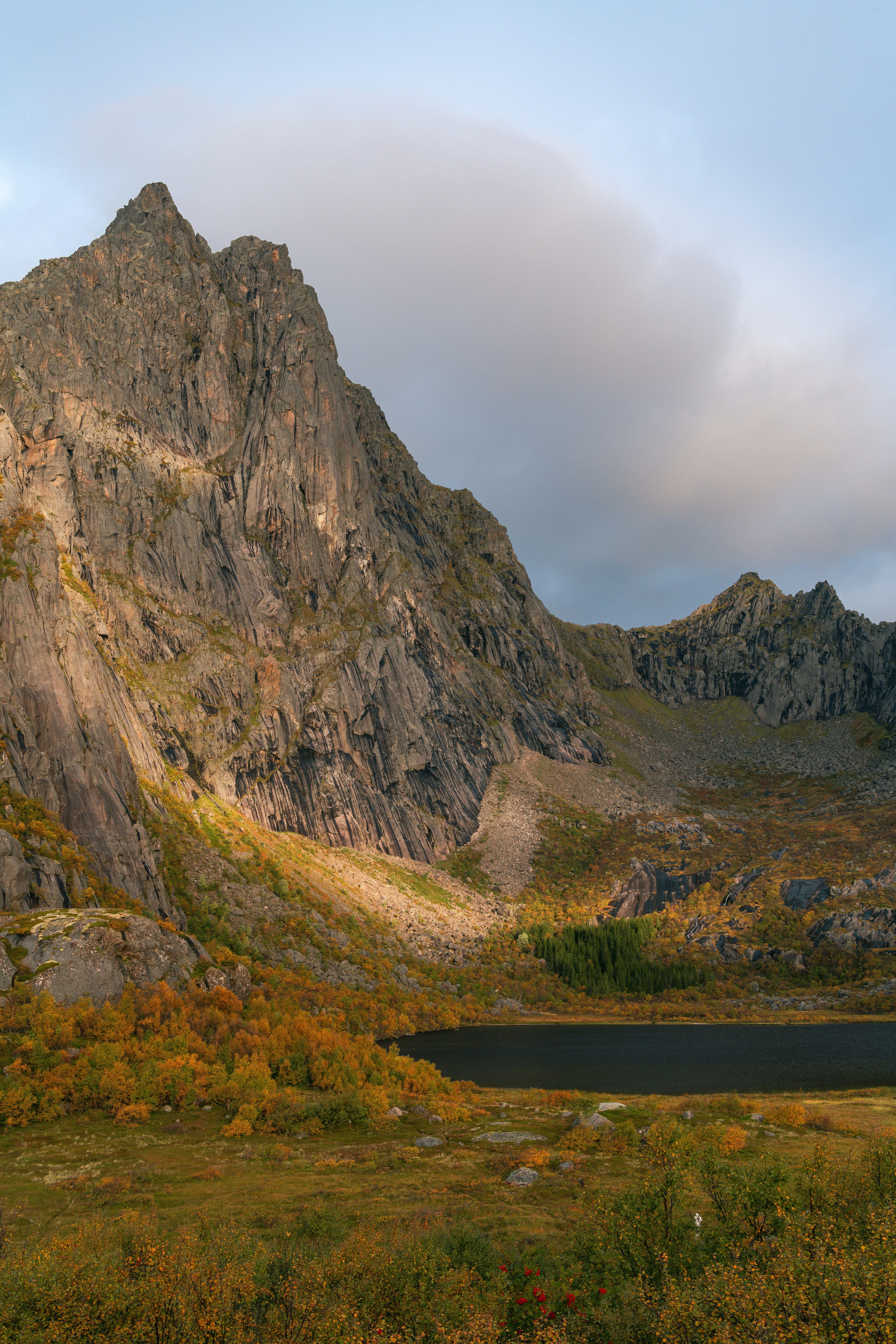 Jagged mountain peak rising above a tranquil alpine lake with autumn foliage in the foreground; a rugged valley scene.