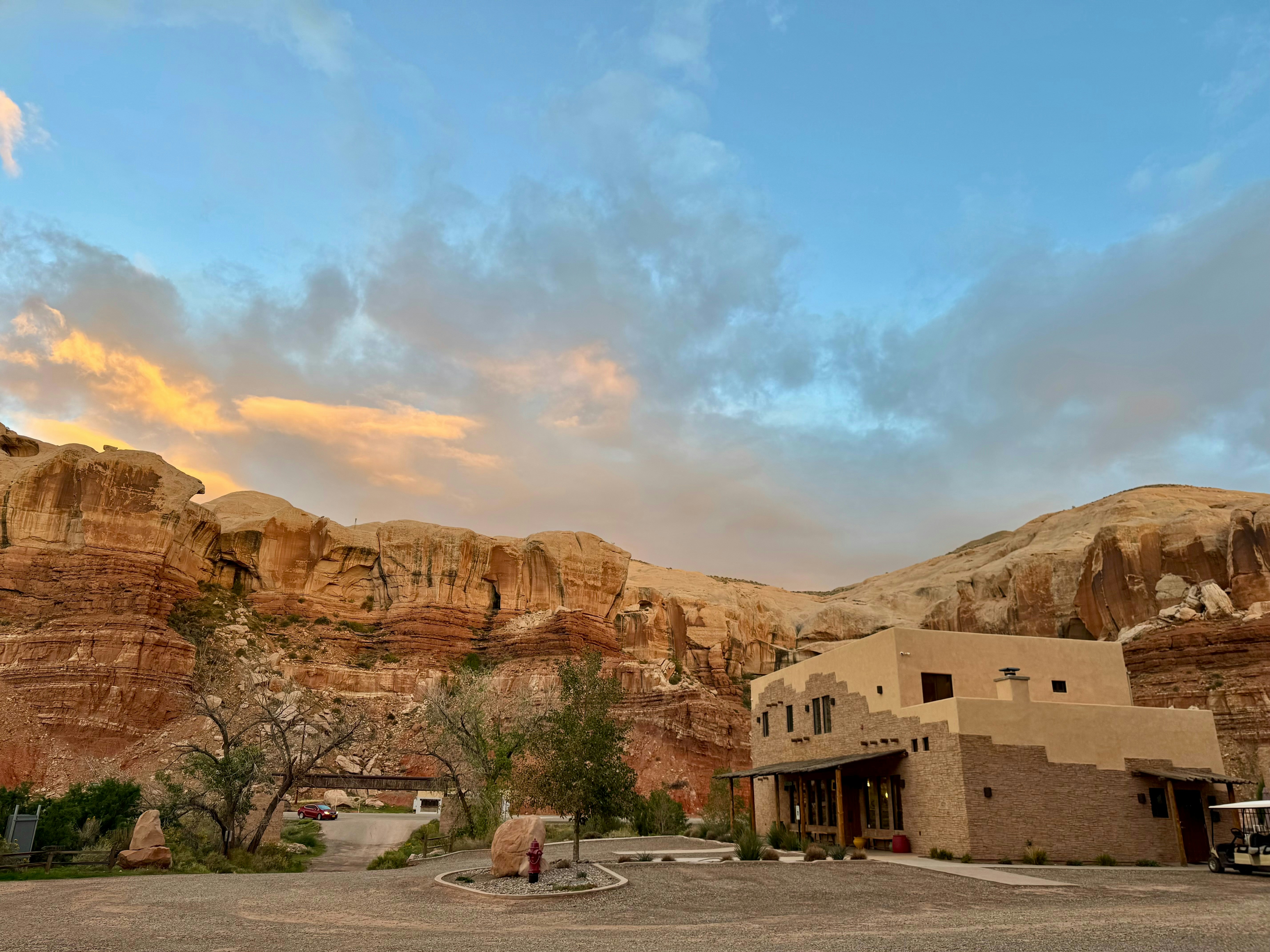A tan adobe building beneath towering red sandstone cliffs under a soft blue and orange sky