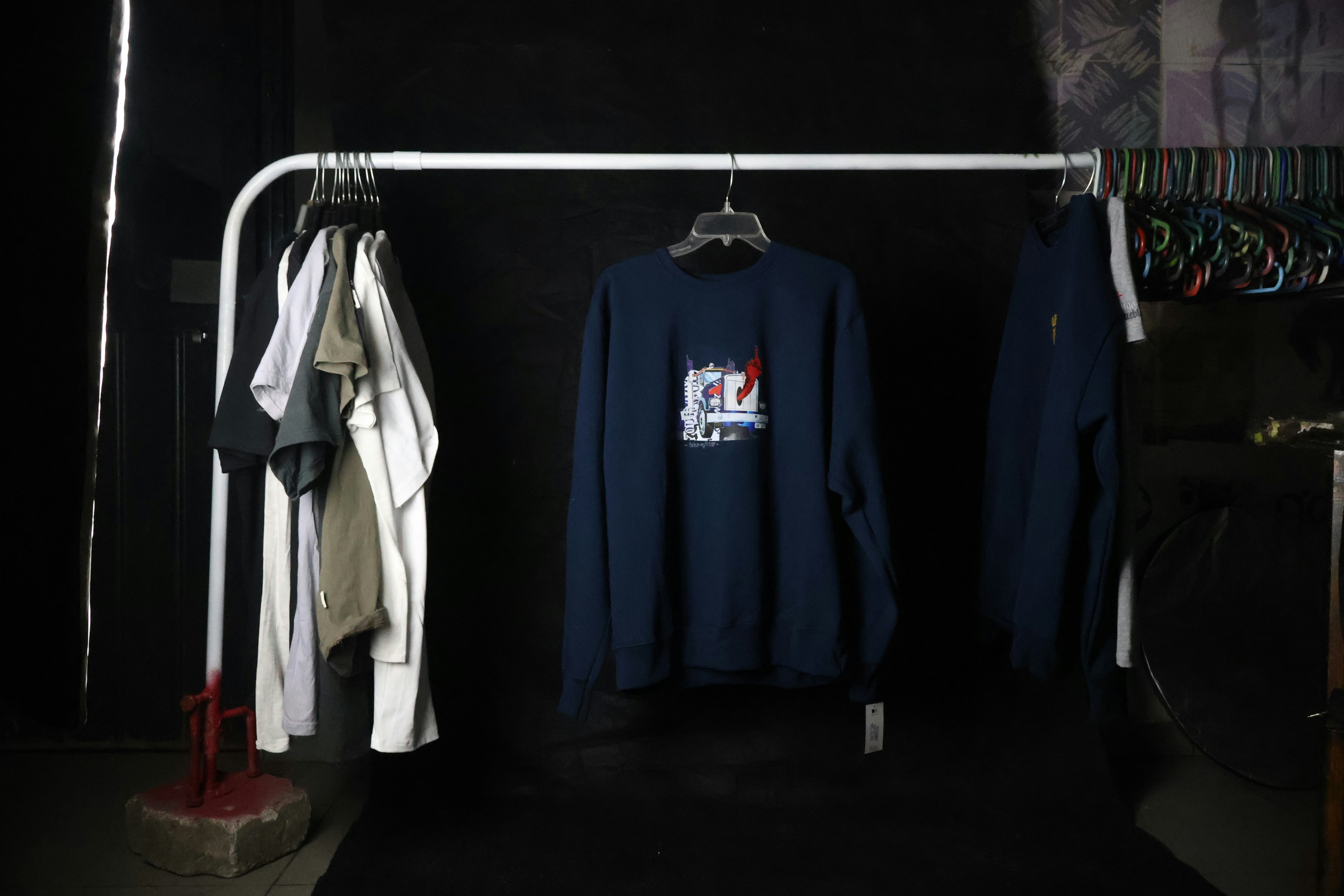 Dark clothing on a metal rack with a single navy hoodie centered against a black backdrop, suggesting a small studio catalog setup.