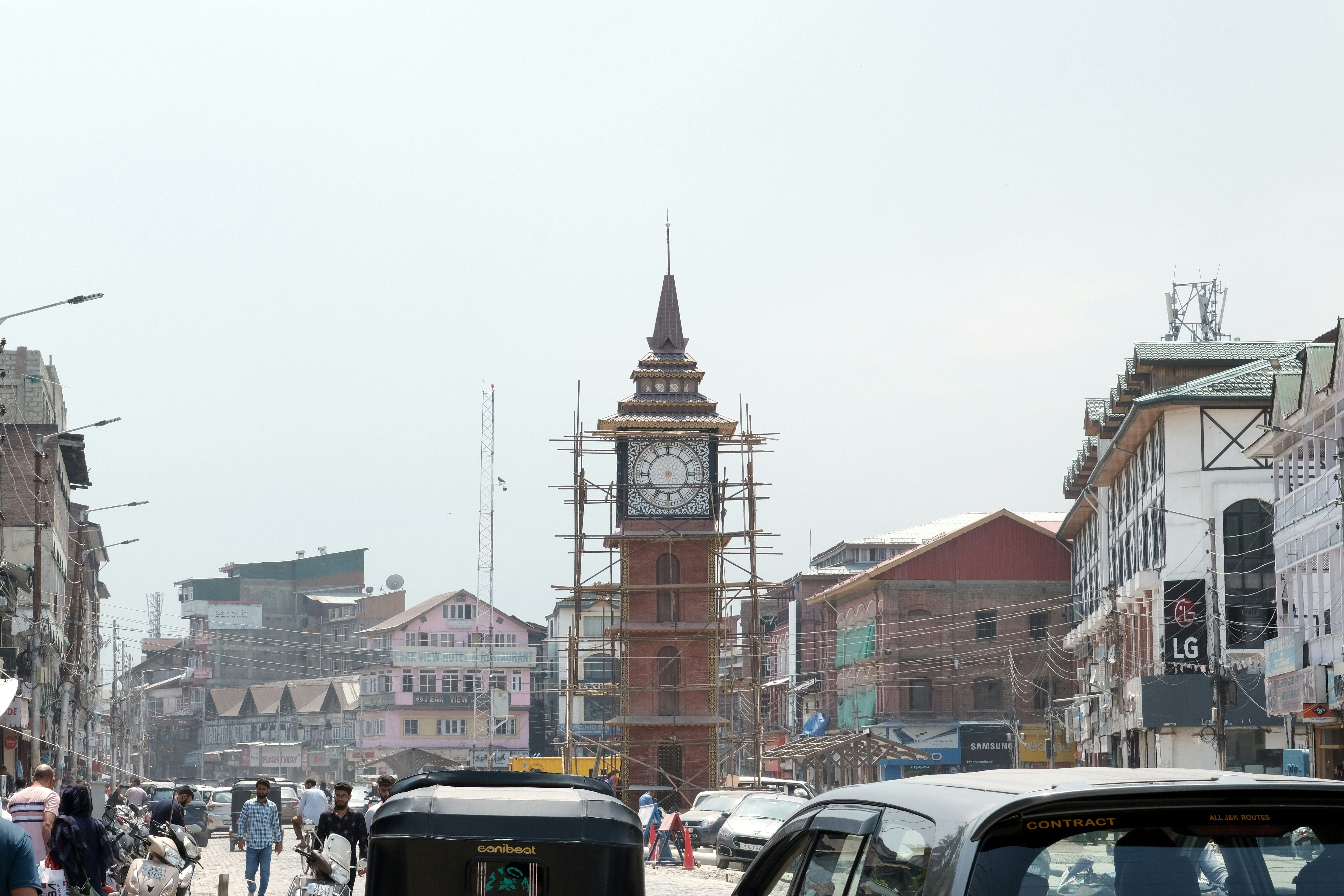 Busy urban street with a prominent clock tower under construction in the center; vehicles and pedestrians fill the foreground, while a row of mixed-era buildings lines the sides. A logo-bearing storefront hints at commercial activity.