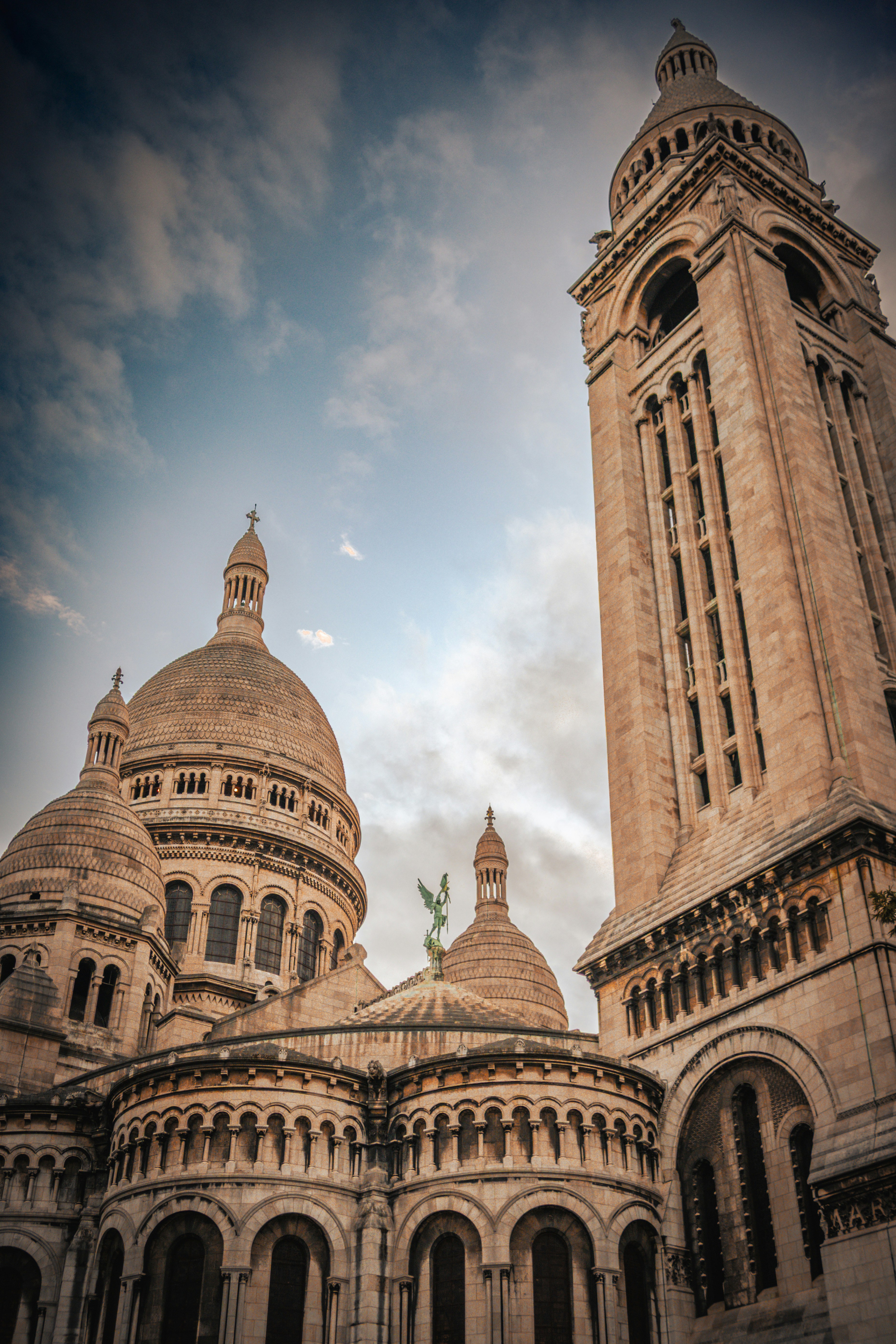 Sculpted domes and a towering spire rise over a stone basilica, with soft sunlight and dramatic clouded sky behind. A statue perched atop the roof adds a subtle focal point.