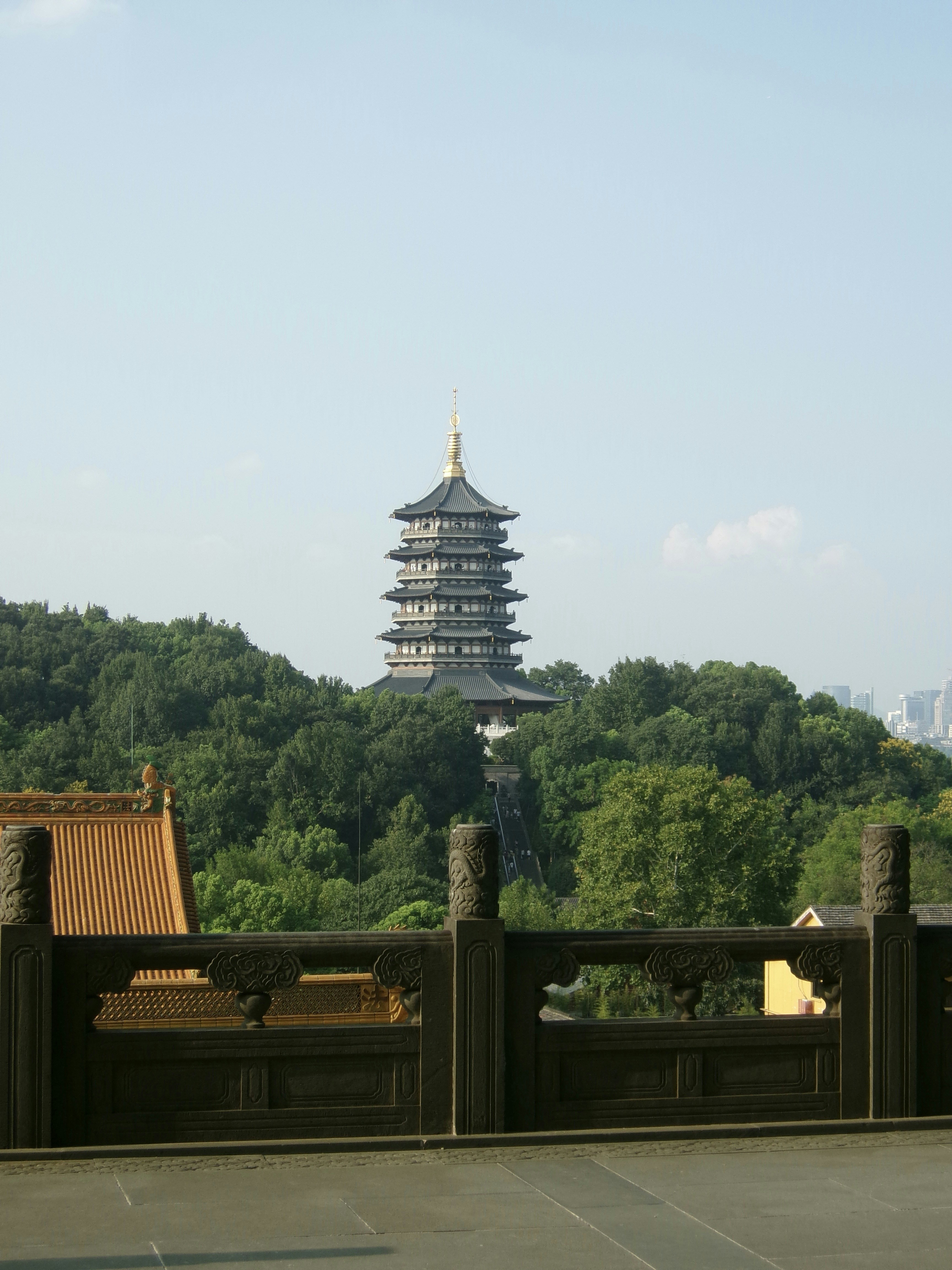 Stone railing foreground with traditional architecture leads to a multi-tier pagoda rising above a treeline under a clear sky.
