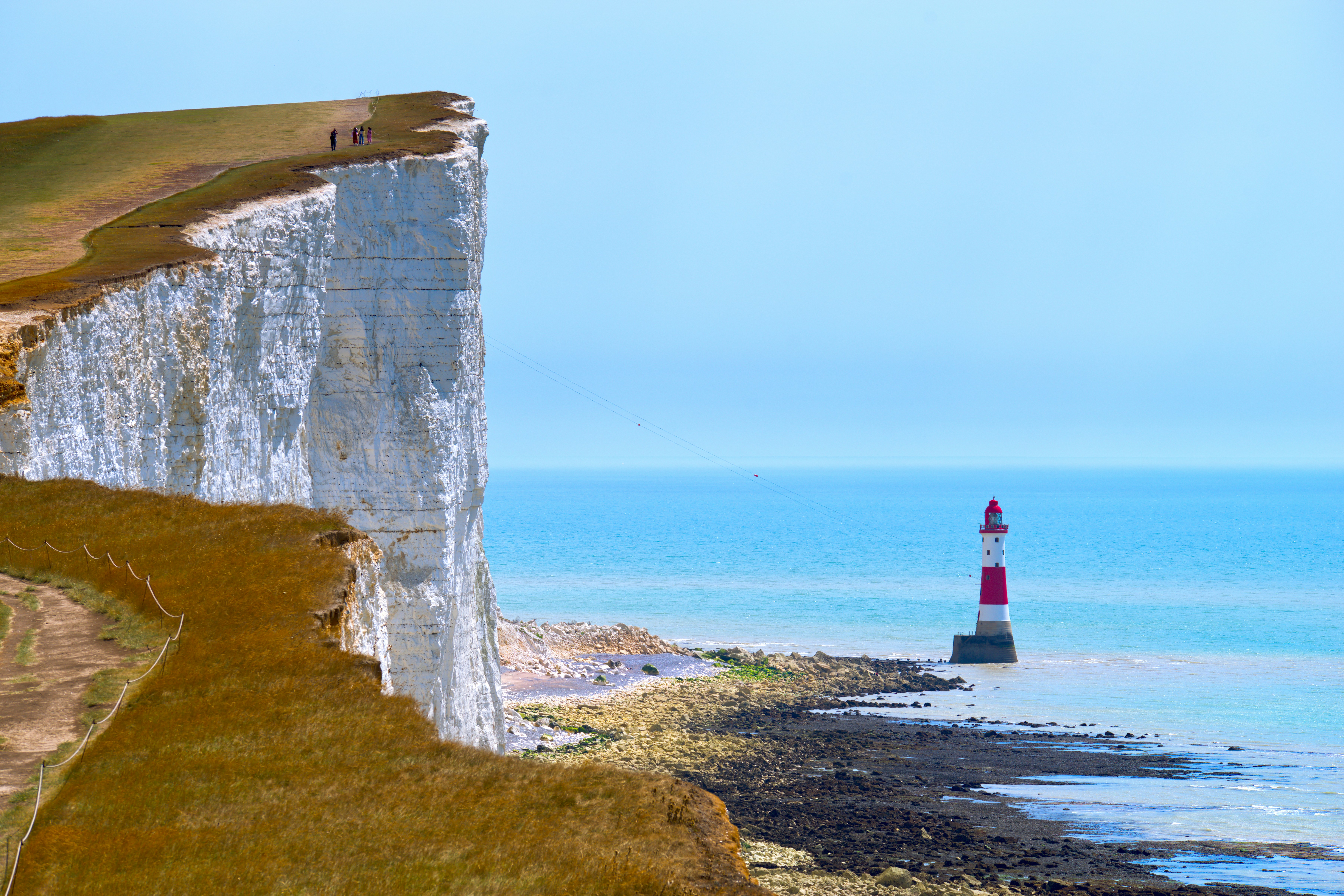 Cliffs drop to a rocky shoreline with a solitary lighthouse standing offshore as calm blue water meets the horizon.