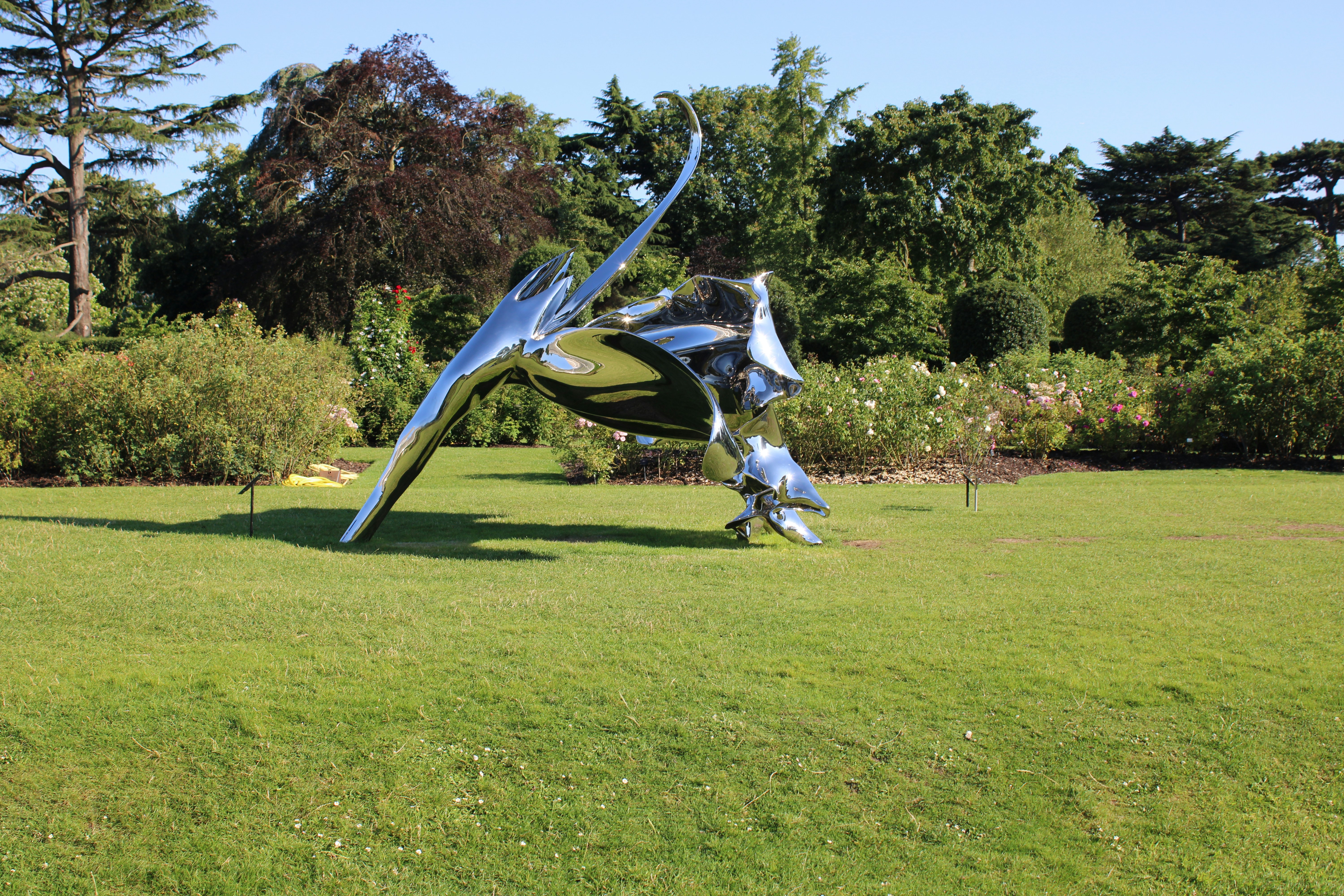 Shiny chrome sculpture of a bird in flight set on a sunlit grassy park with trees in the background.
