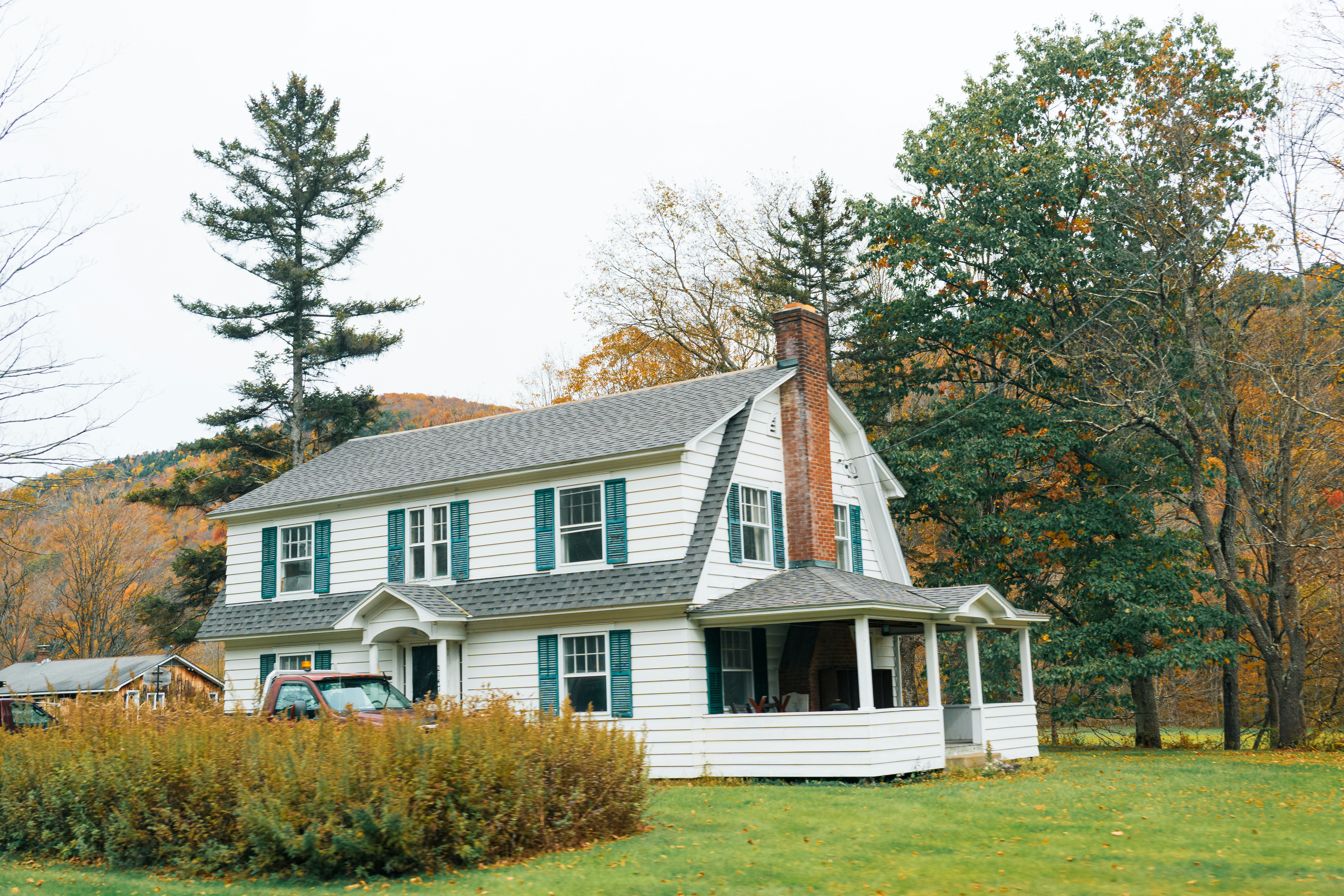 White two-story house with blue shutters in a yard of green grass and fall foliage, featuring a wraparound porch and chimney, captured in soft daylight.