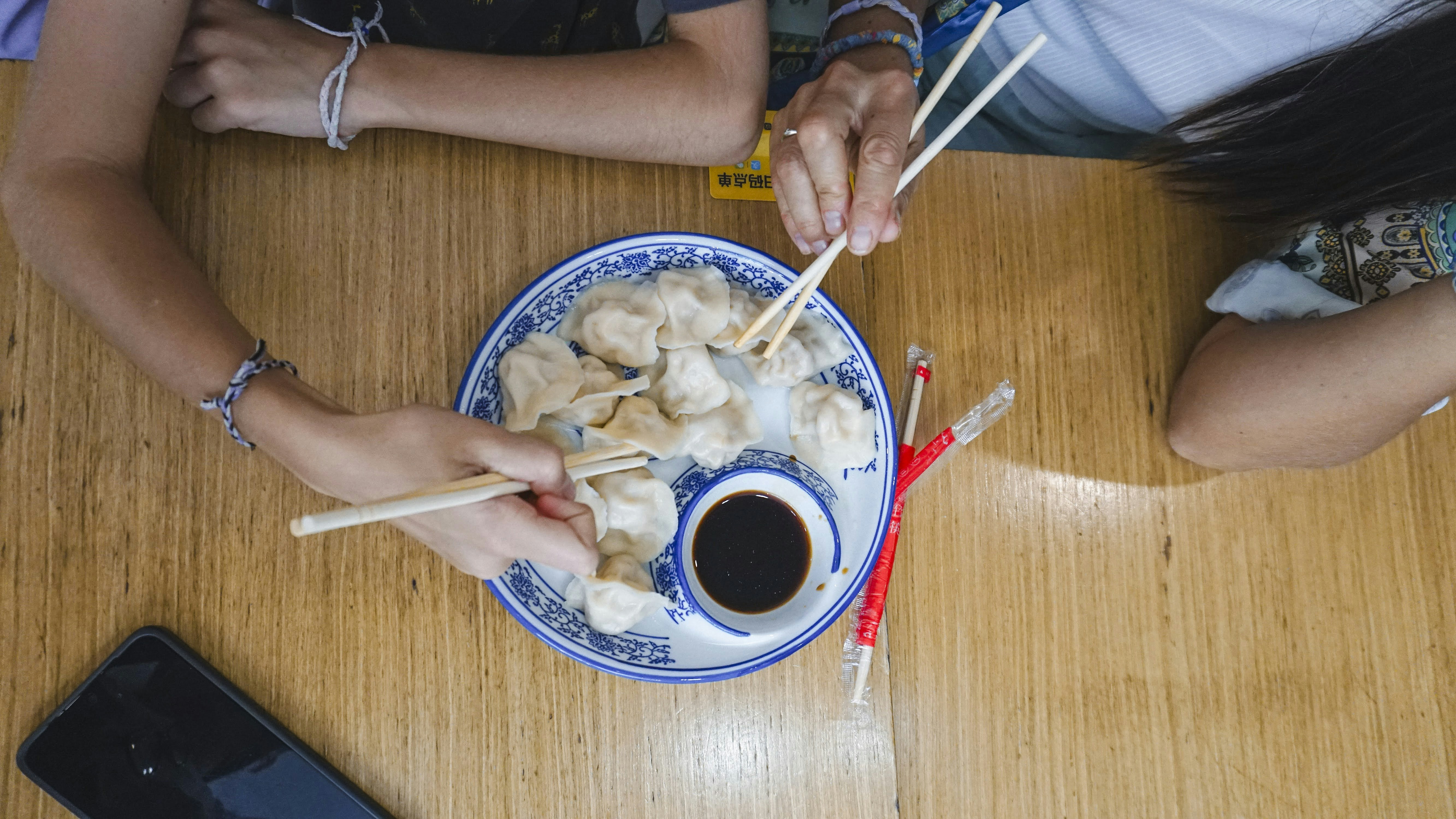 Plate of dumplings with soy sauce and chopsticks at a wooden table; hands reach in from both sides, hinting at a shared meal. Casual dining scene with a phone nearby and colorful chopstick wrappers.