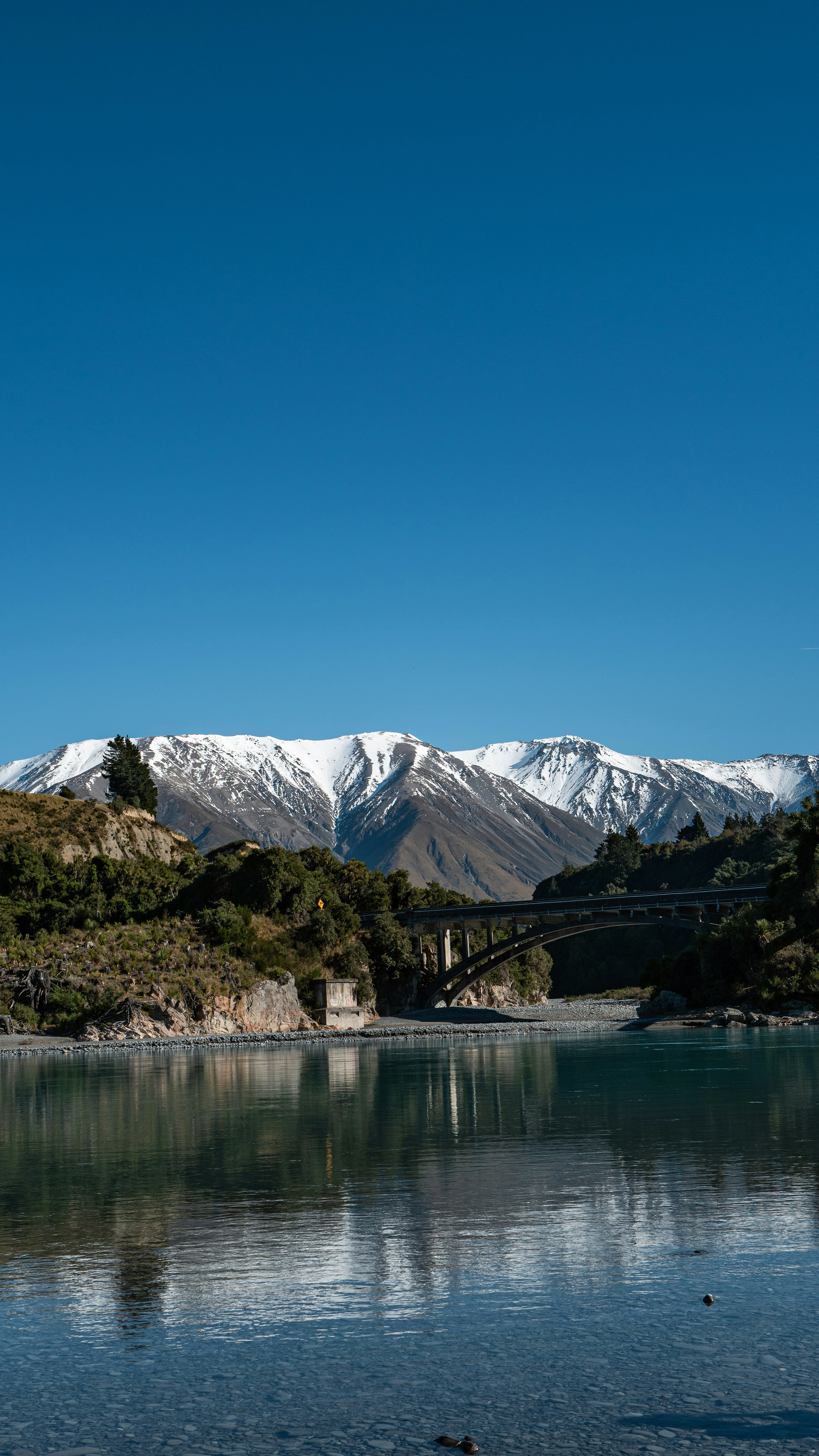 Snow-dusted mountains rise above a calm river, with a bridge weaving through the midground and evergreen trees lining the banks.