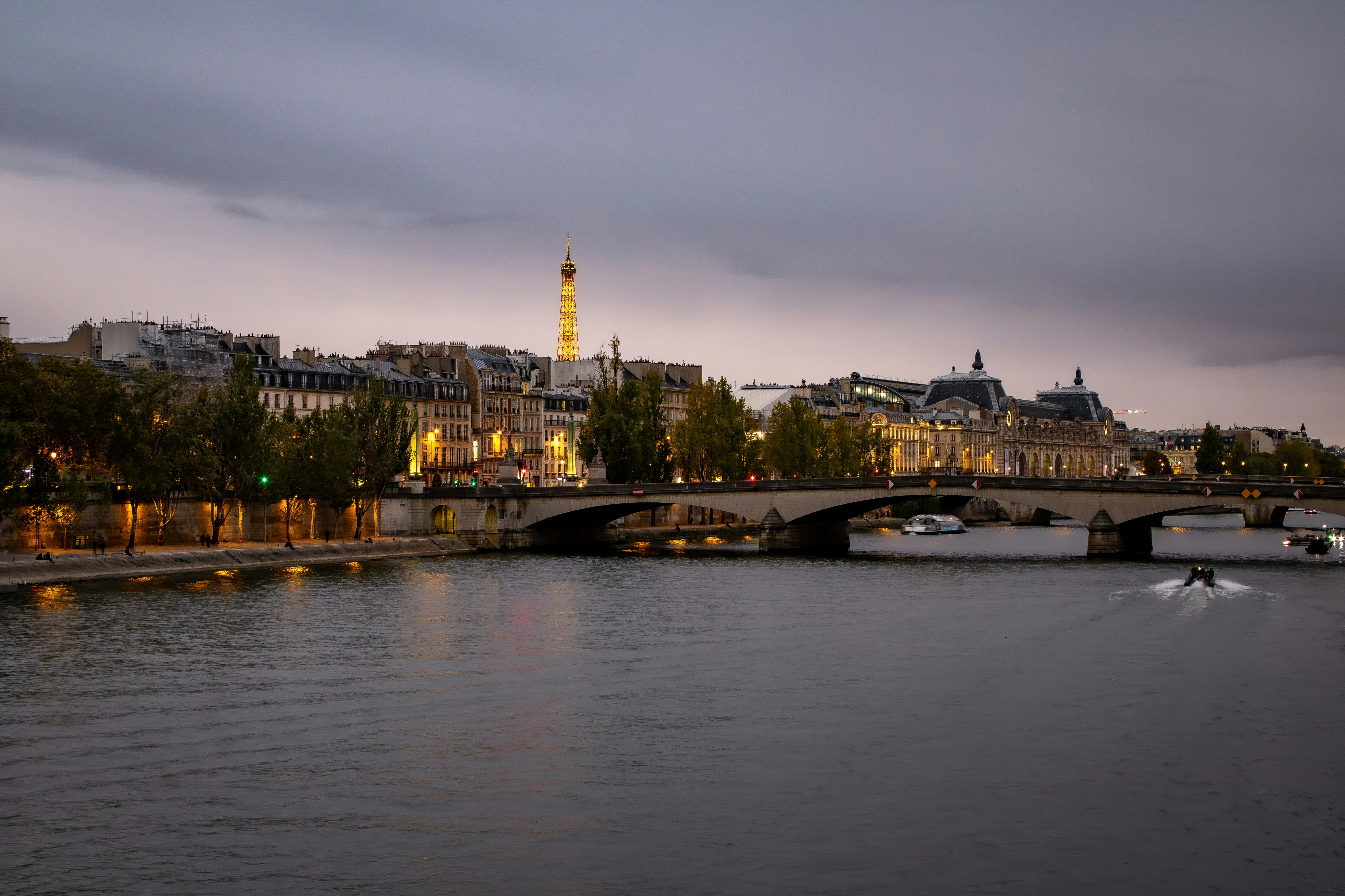 Evening view of a historic riverside with illuminated buildings and a bridge over calm water along the Seine. The scene captures a skyline with a tall lit tower and a small speedboat trail, emphasizing ambience and architecture.