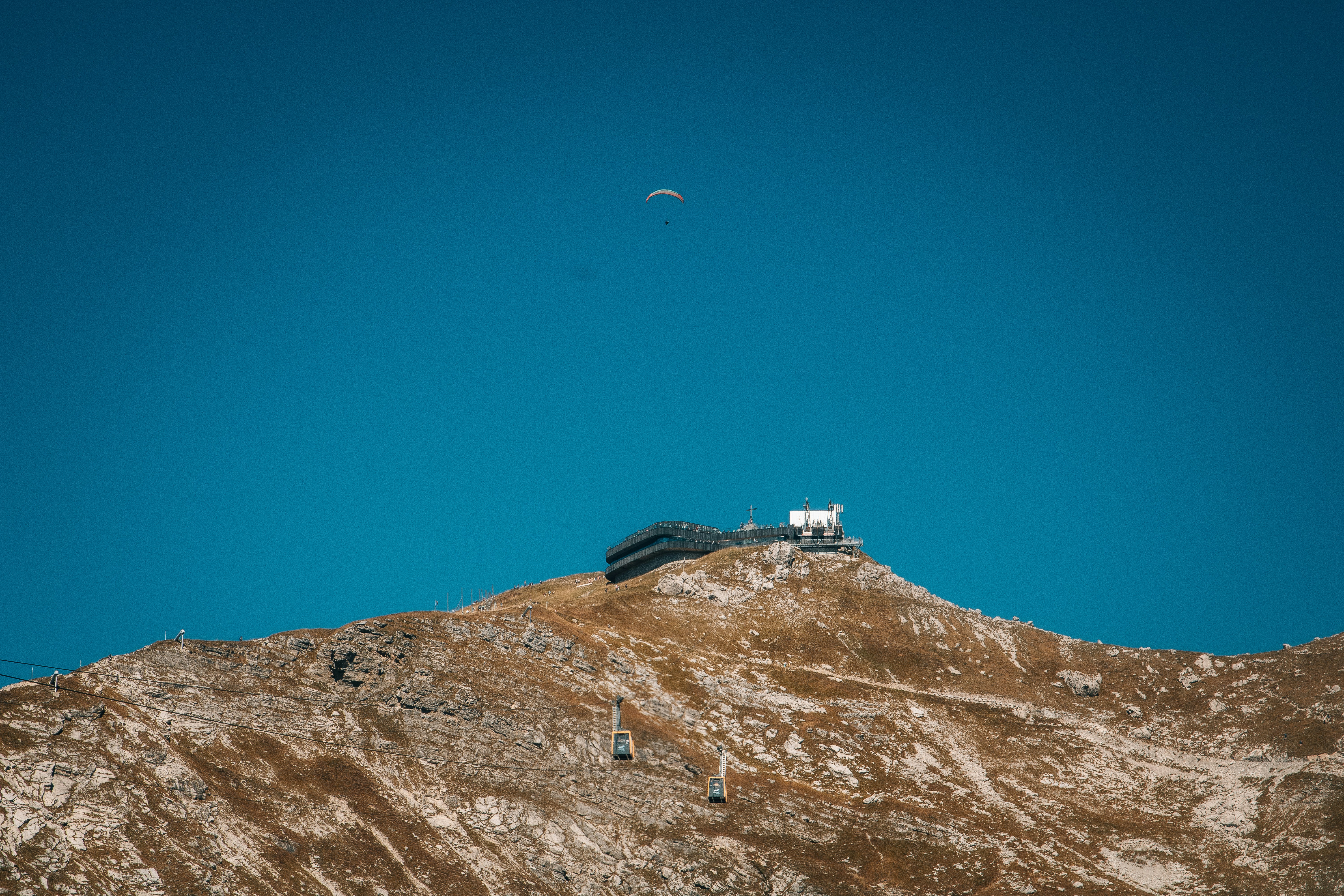 Rugged mountain ridge with a small building at the summit under a clear blue sky, featuring ski-lift cables in the slope foreground.