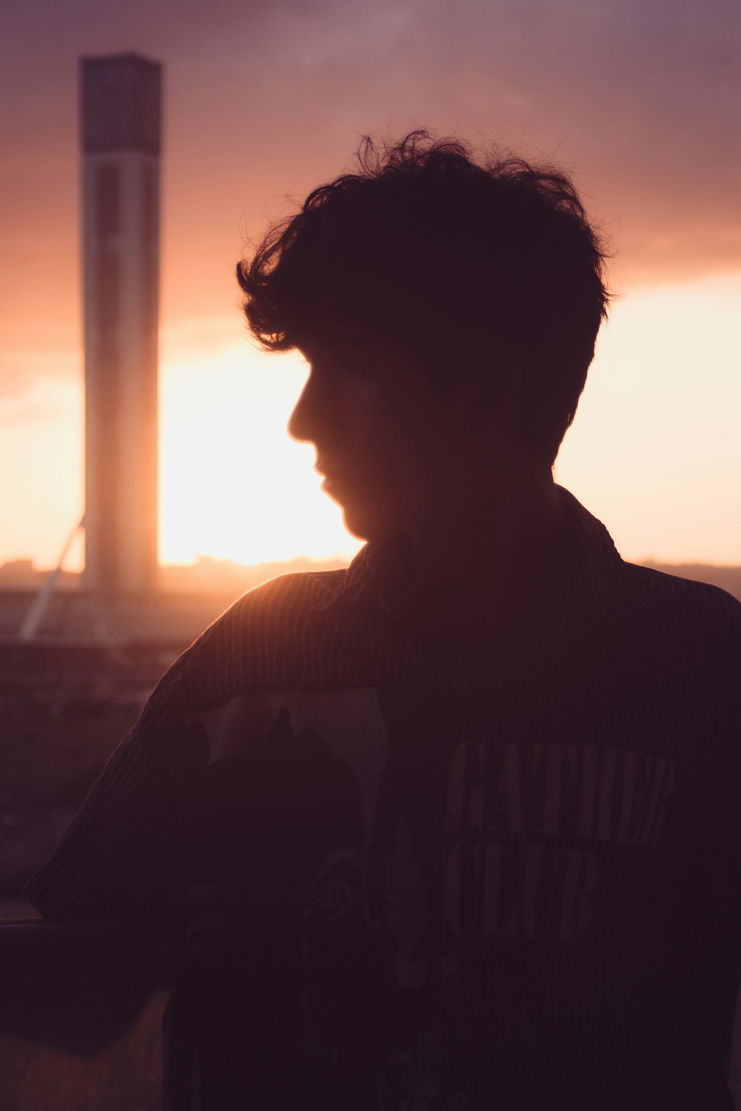 Silhouetted profile of a person against a warm sunset, with an industrial tower in the background; the scene emphasizes backlit mood and shape over detail. This is a photographic portrait.