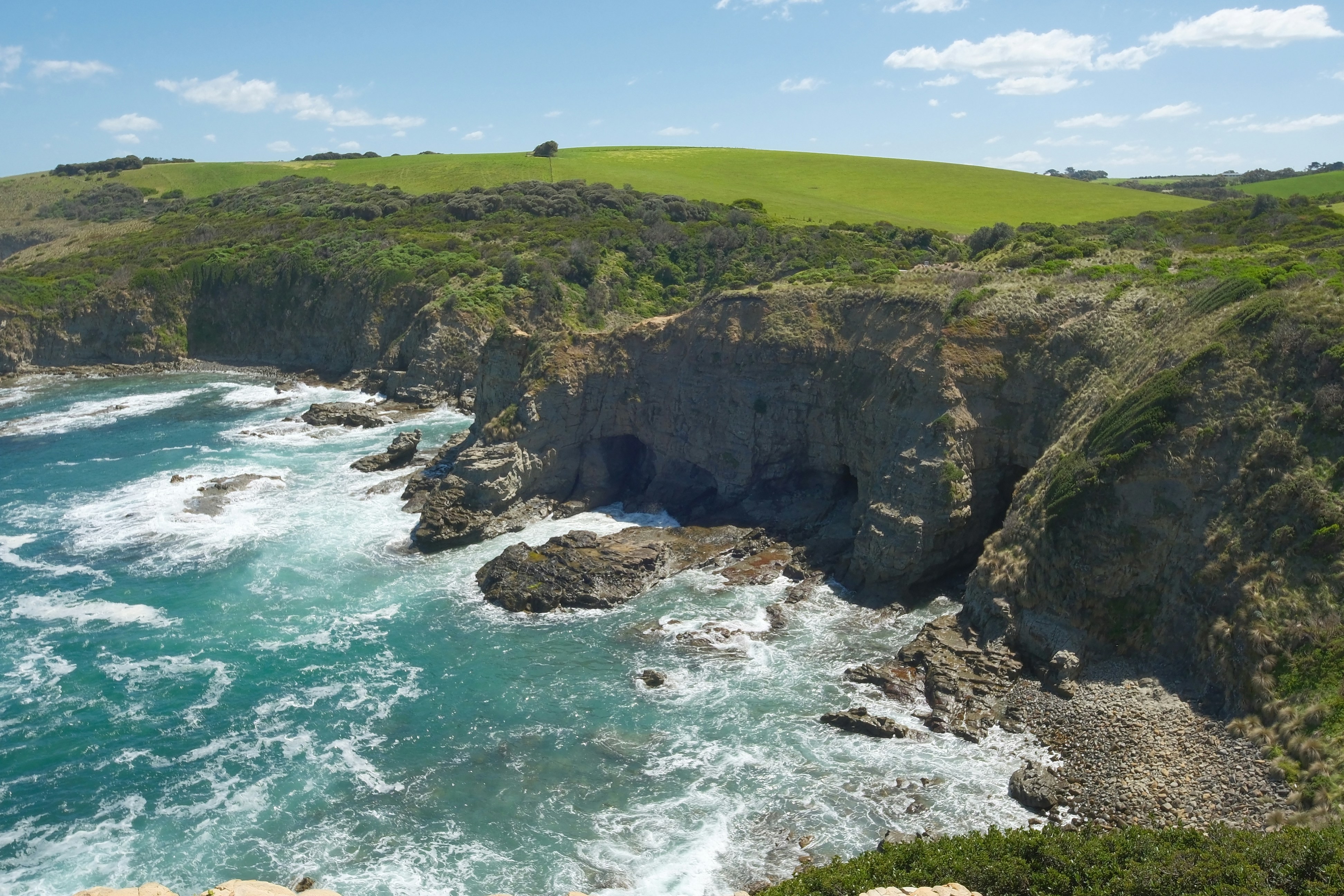 Sunlit coastal cliffs with turquoise waves crash against rocky outcrops and a grassy headland; a panoramic seascape highlighting rugged geology and vivid surf.