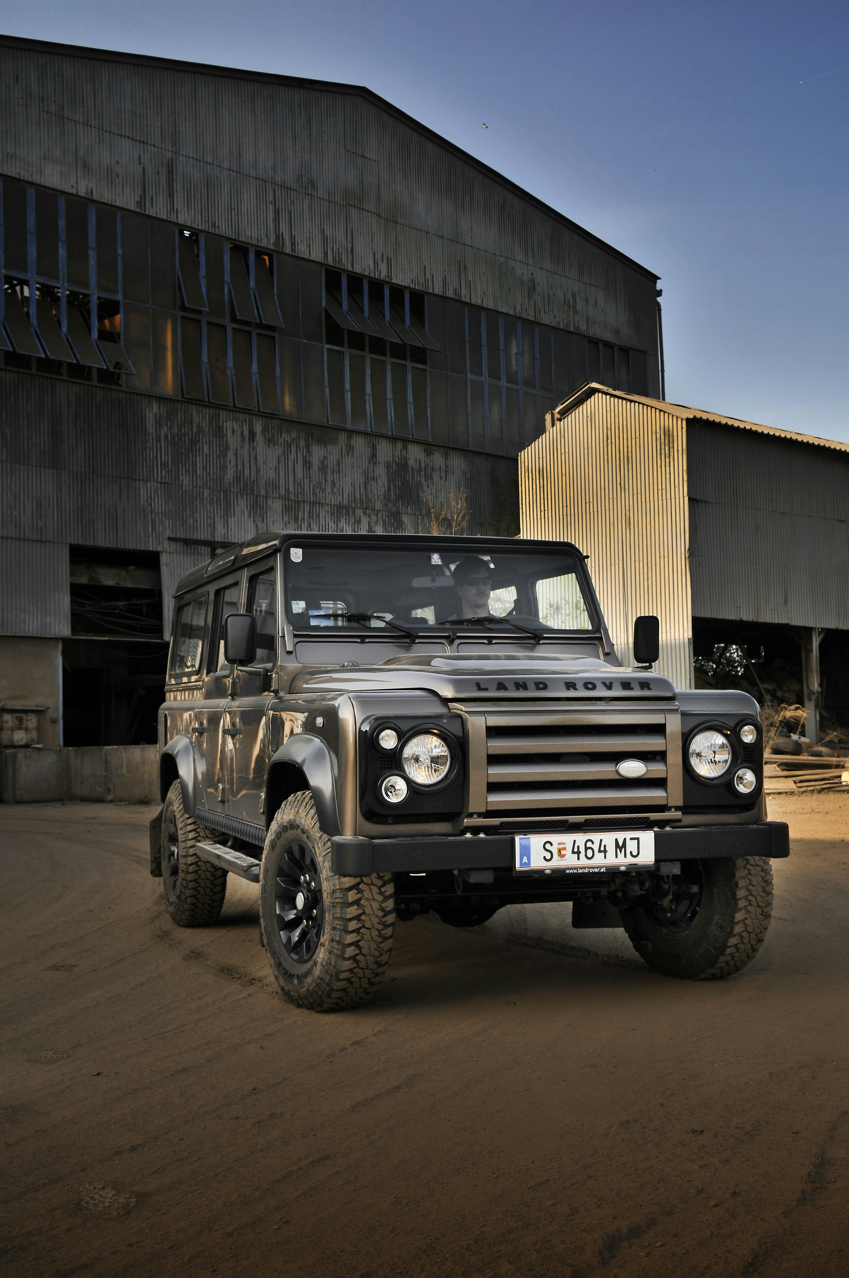 A dark gray Land Rover Defender parked in front of a weathered industrial warehouse, sunlight casting long shadows on the dusty ground. The scene focuses on the sturdy vehicle and its rugged stance amid aged corrugated structures.