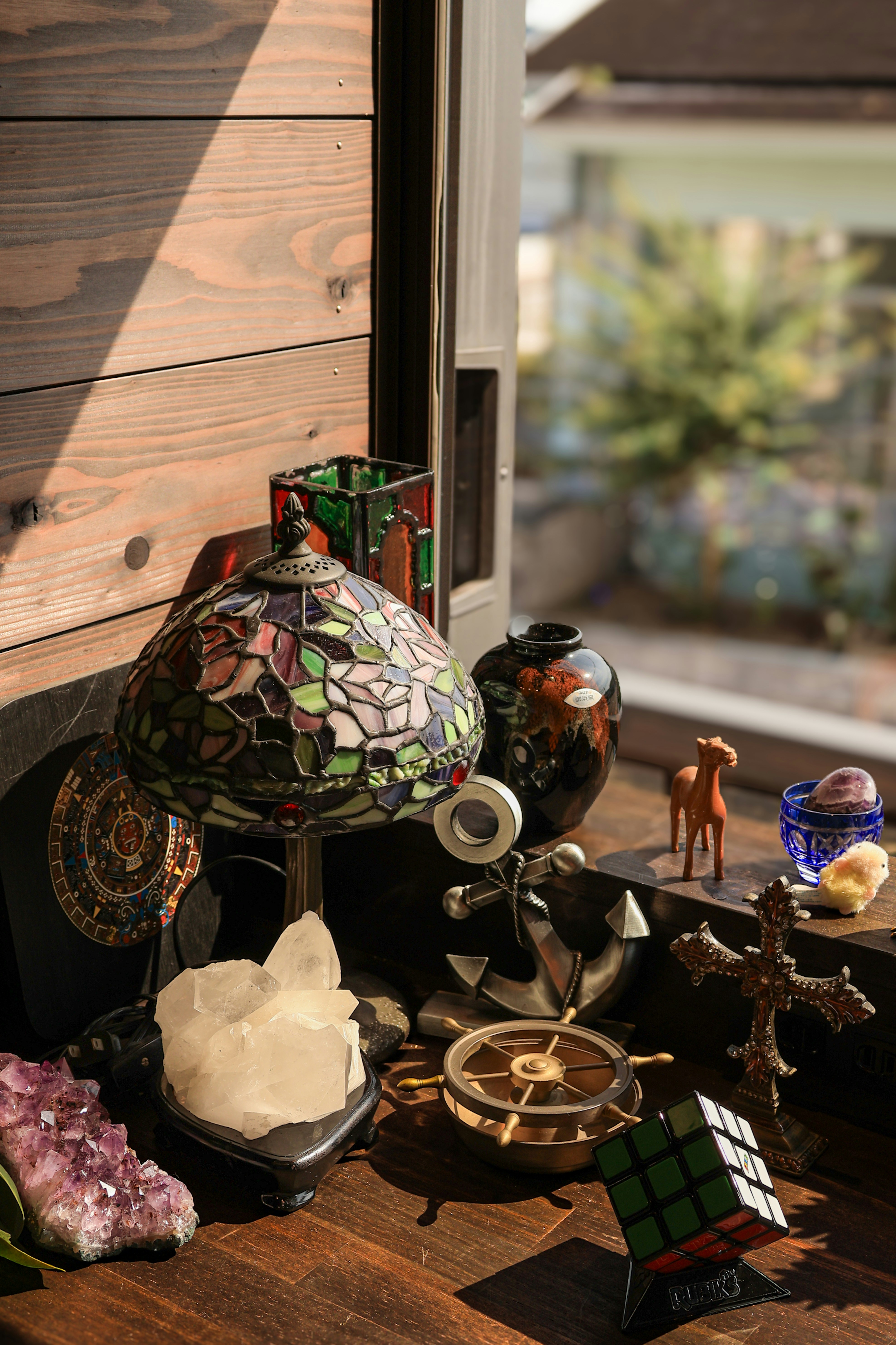 Decorative mosaic lamp and assorted curios sit on a wooden surface near a sunlit doorway. The arrangement includes crystals, a Rubik's cube, a compass-like device, and small figurines with a blurred garden background.