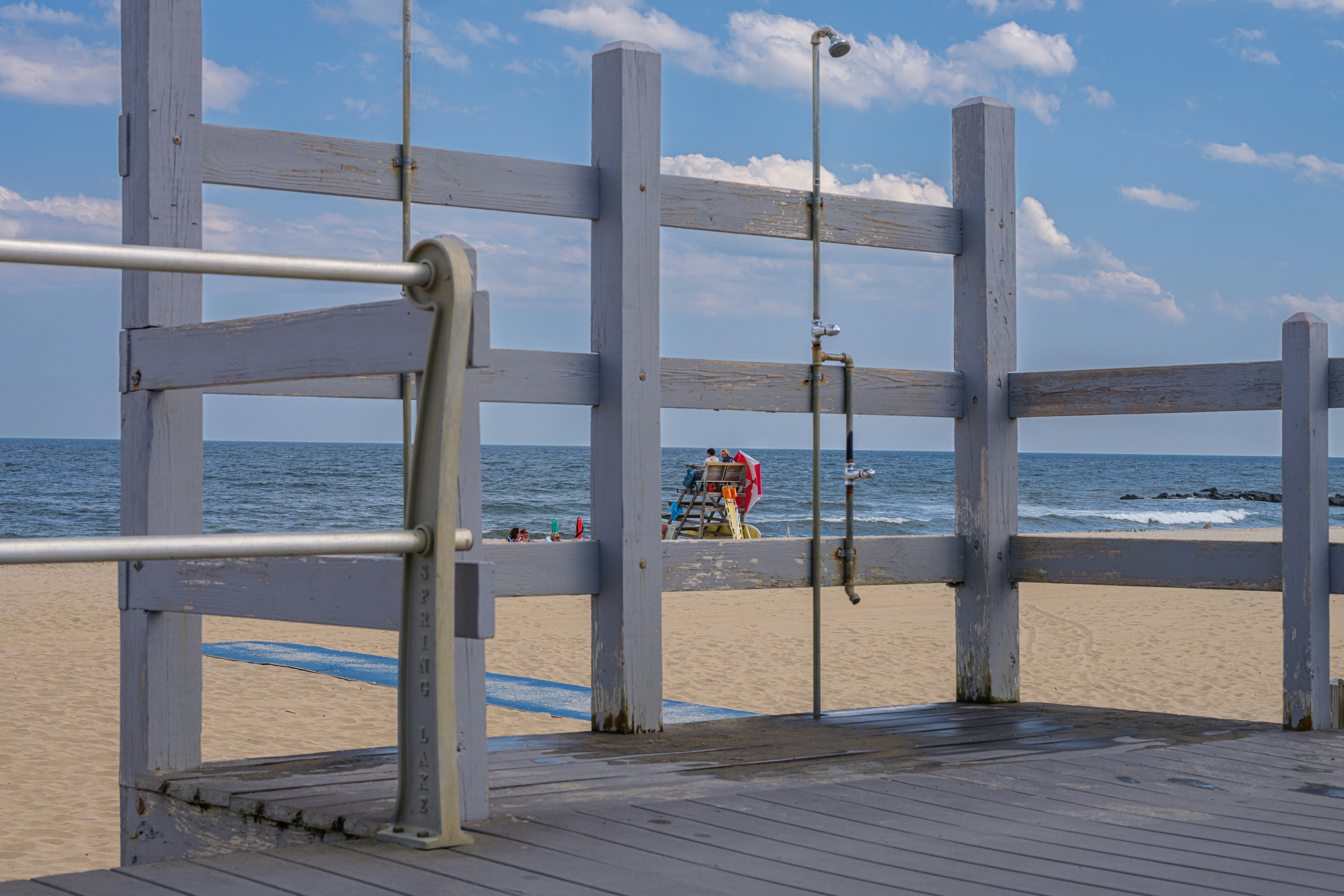 Lifeguard stand visible beyond a wooden railing on a sunlit beach, with calm sea and scattered people in the distance.