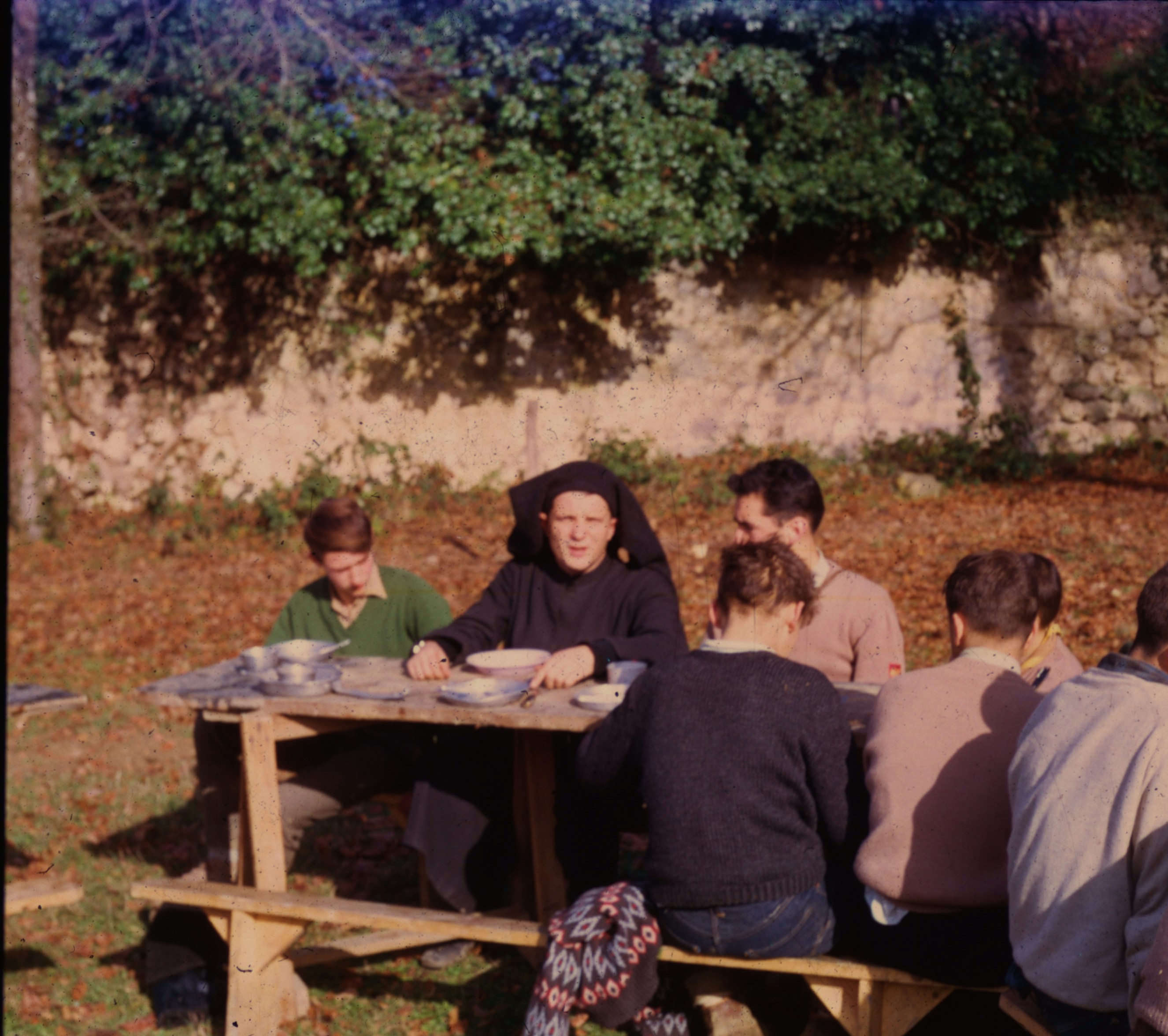 Outdoor communal meal at a sunlit picnic table, faces gathered around plates