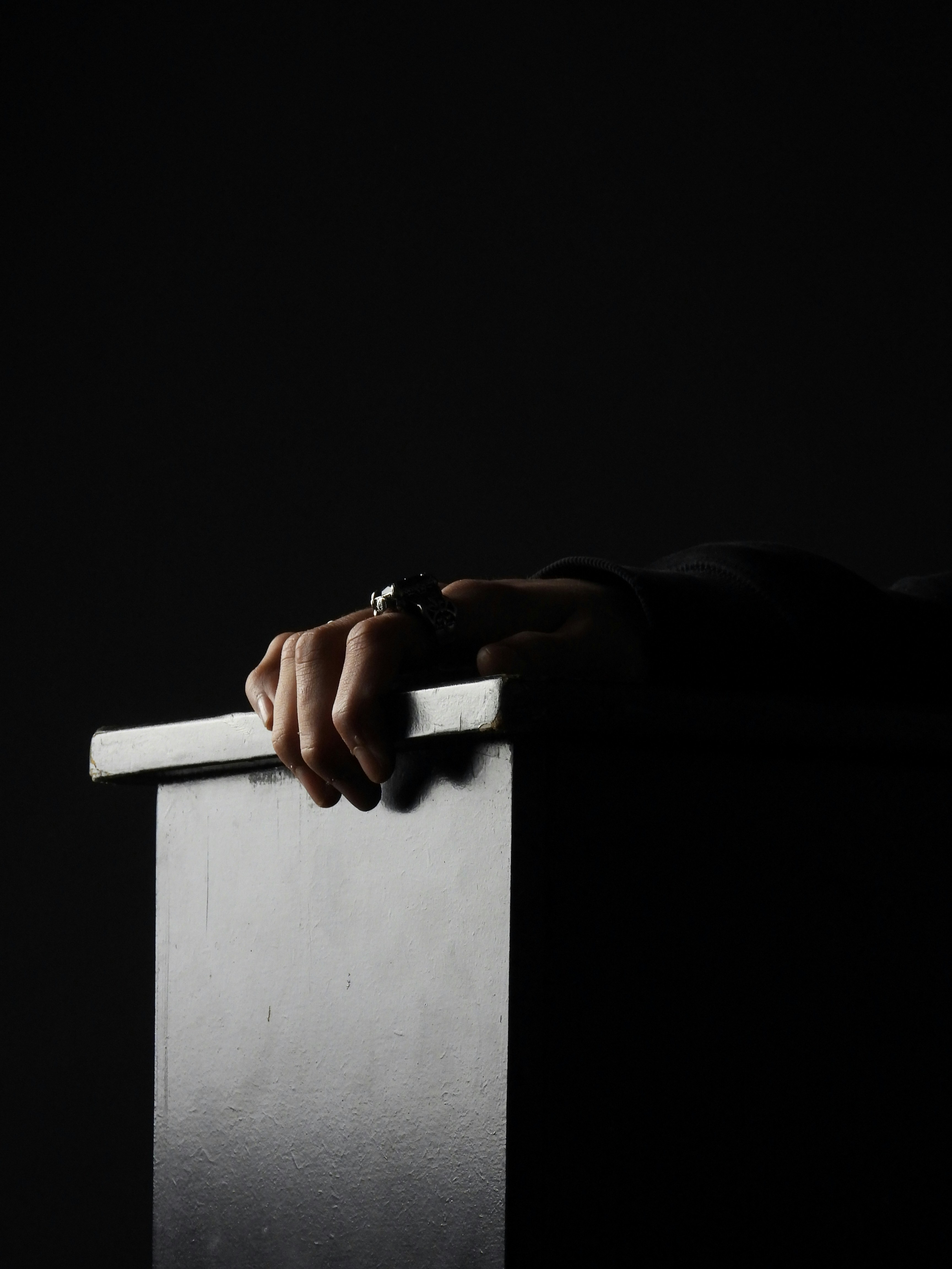 Dark, minimalist portrait focusing on a lone hand gripping a ledge, rendered with stark contrast in a nearly black studio backdrop. The composition emphasizes texture and subtle metallic gleam on the surface.