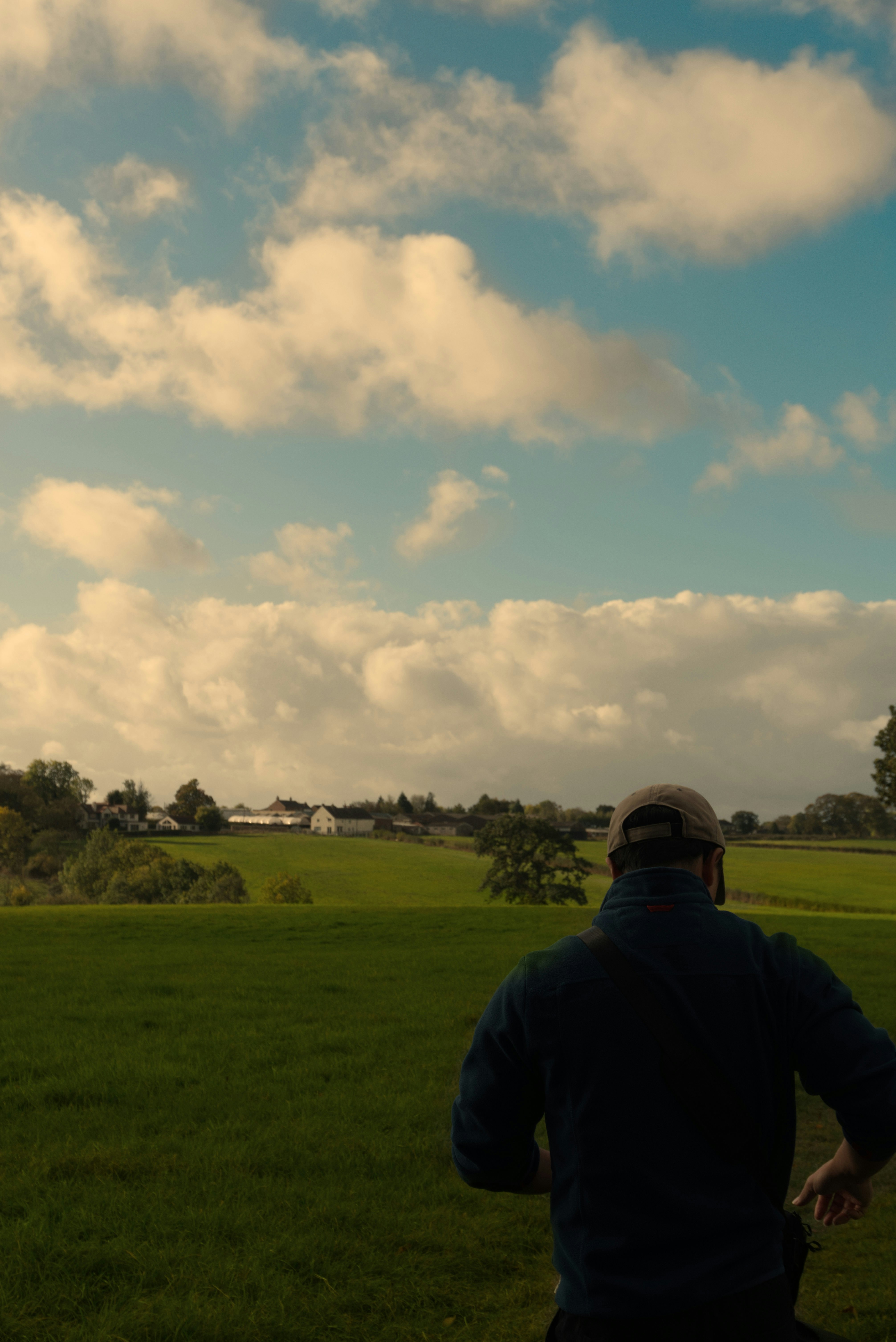 A lone person in a dark jacket runs across a sunlit countryside, with rolling green fields and a distant cluster of houses beneath a partly cloudy sky.