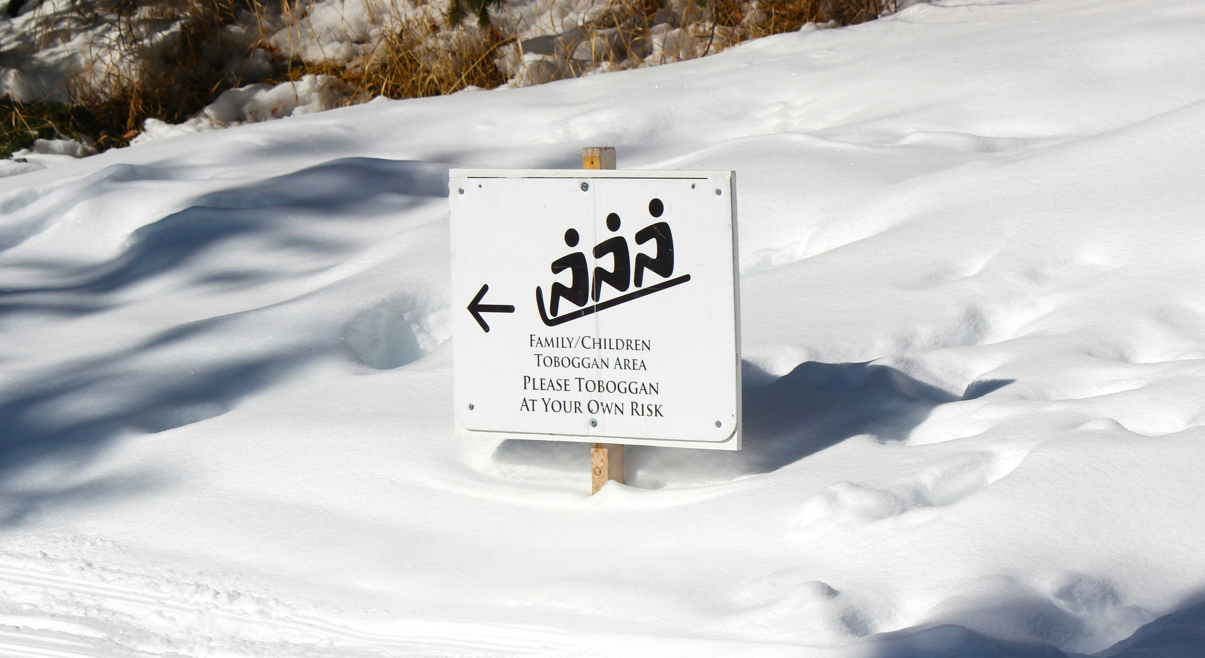 Sign warning of tobogganing risk sits in deep snow; warning text visible amid bright, sunlit snowdrifts.