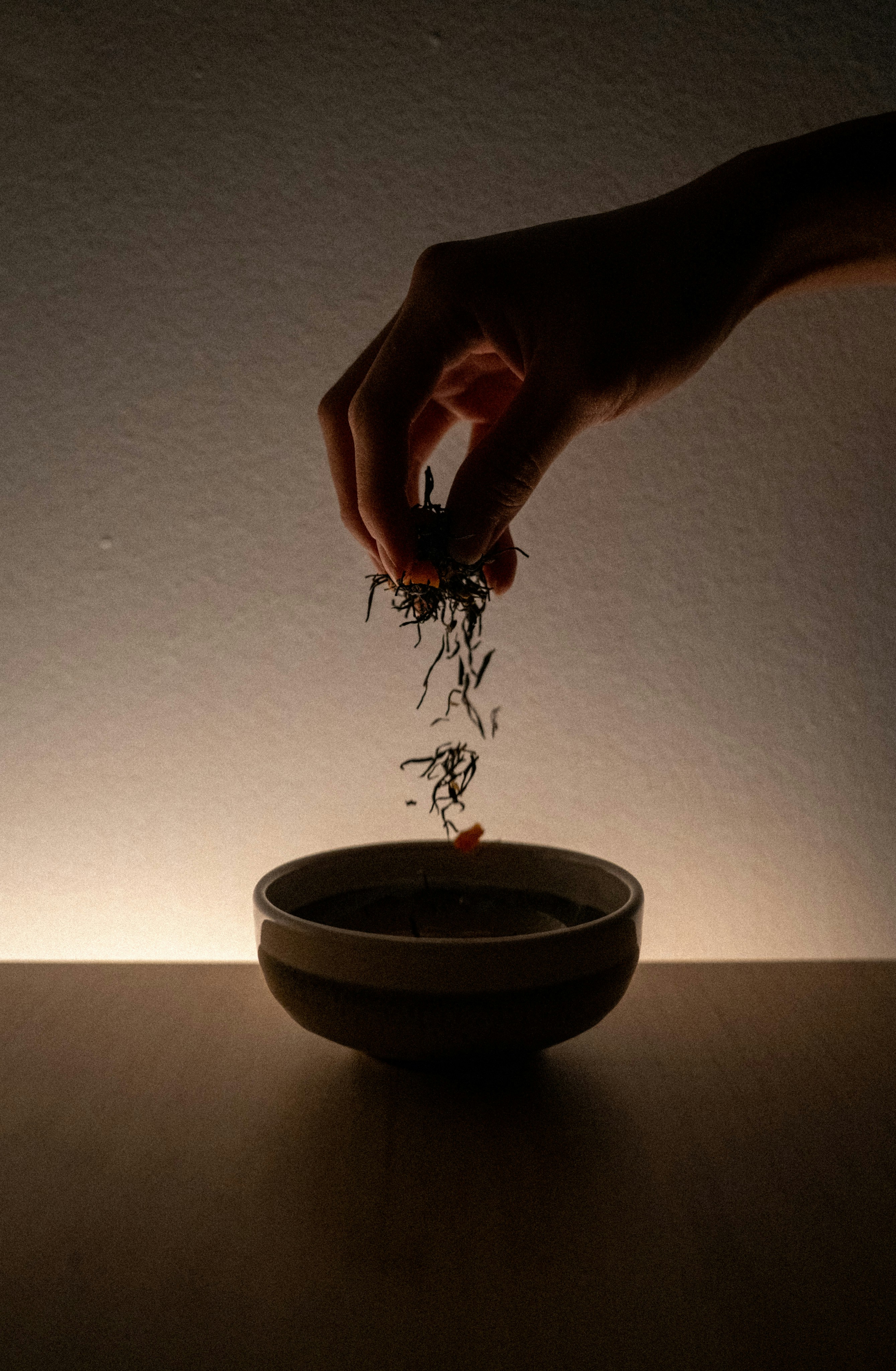A single hand releases wispy fragments toward a shallow bowl against a softly lit backdrop. The composition emphasizes balance between material and emptiness, with the fragments dancing as they fall.