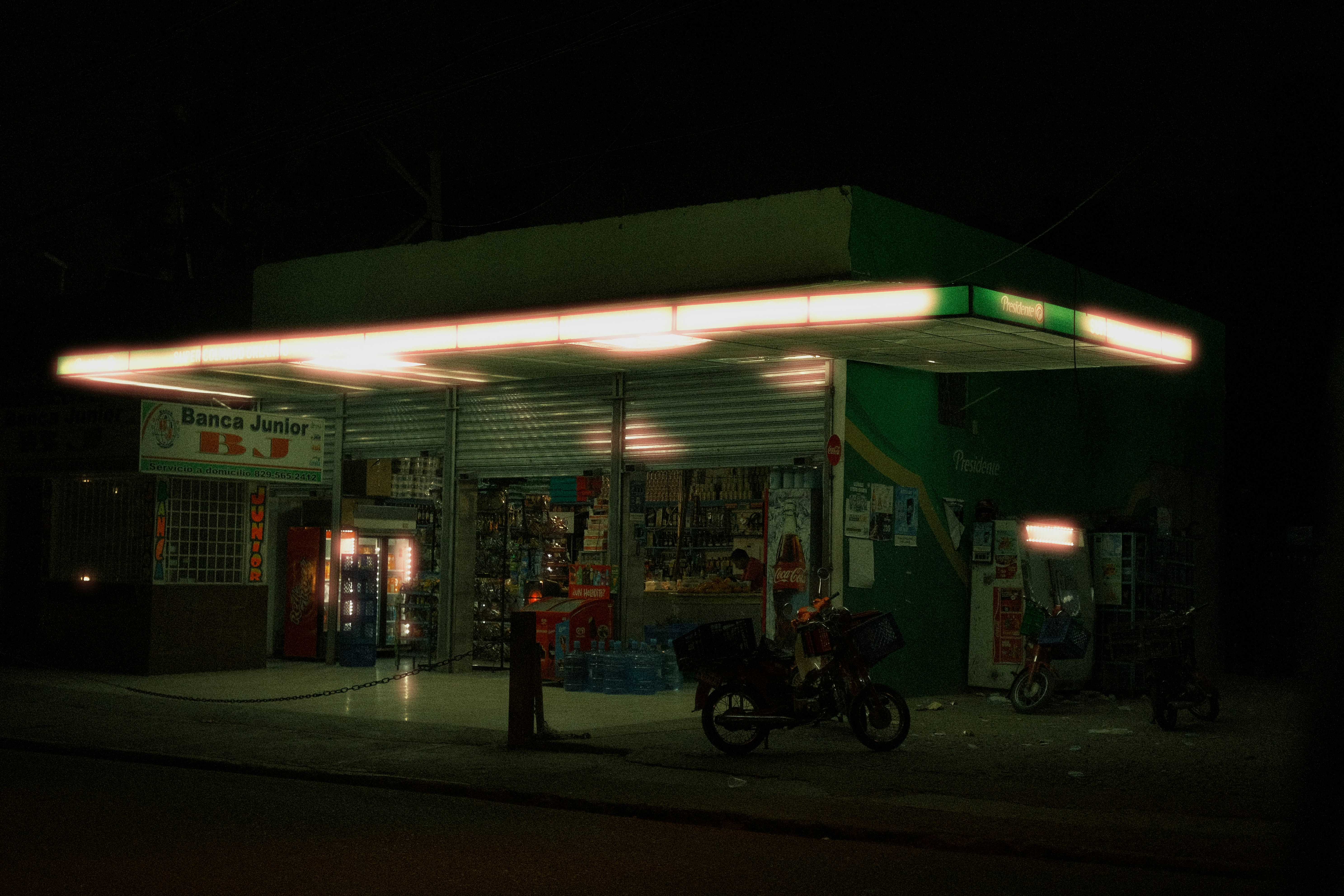 Nighttime storefront with illuminated canopy and a small shopfront; a scooter sits outside while vending machines glow, capturing a quiet street scene at night.