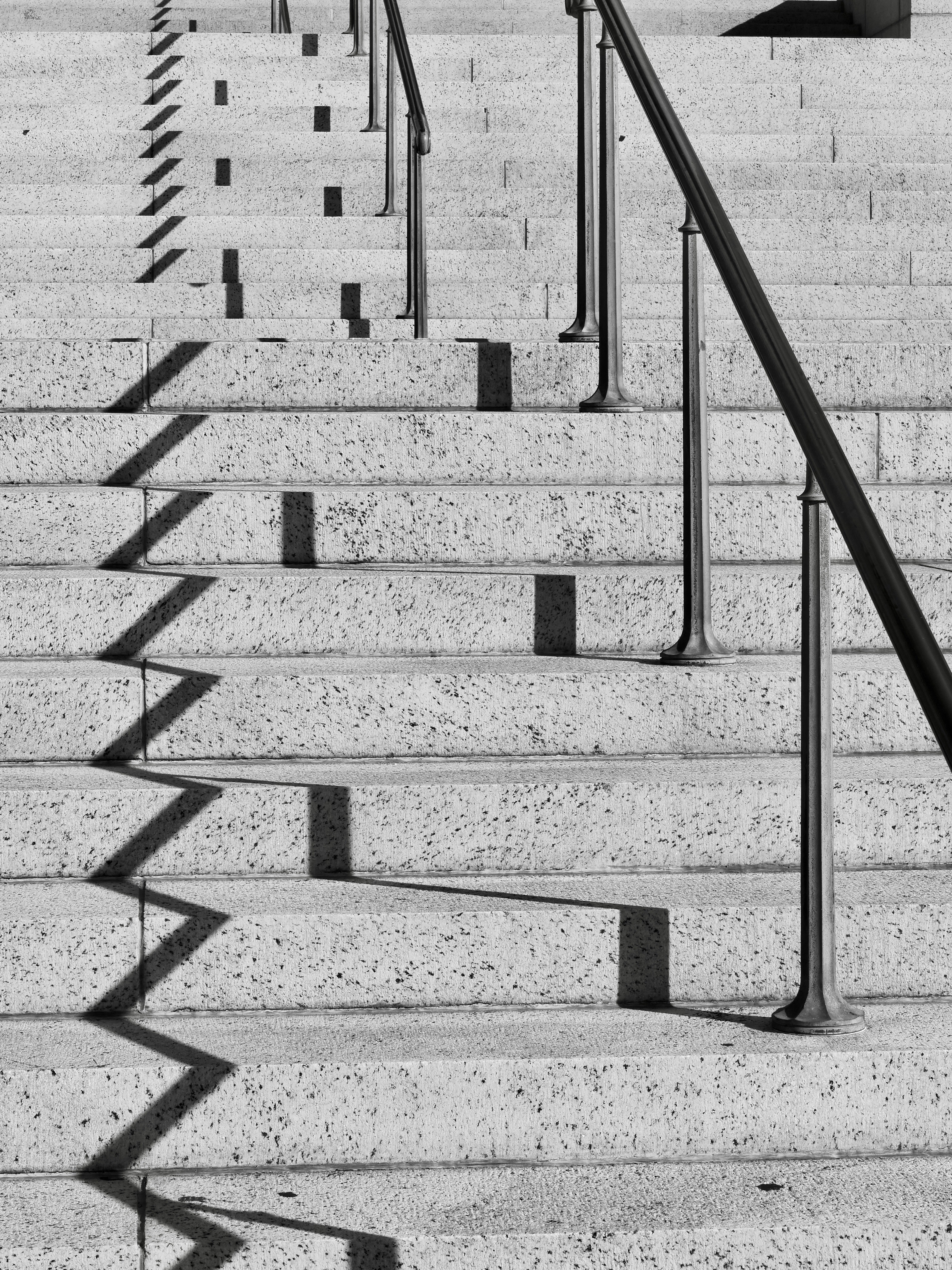Concrete staircase with alternating shadows and metal handrails forming angular patterns; stark monochrome composition emphasizing geometry and contrast.