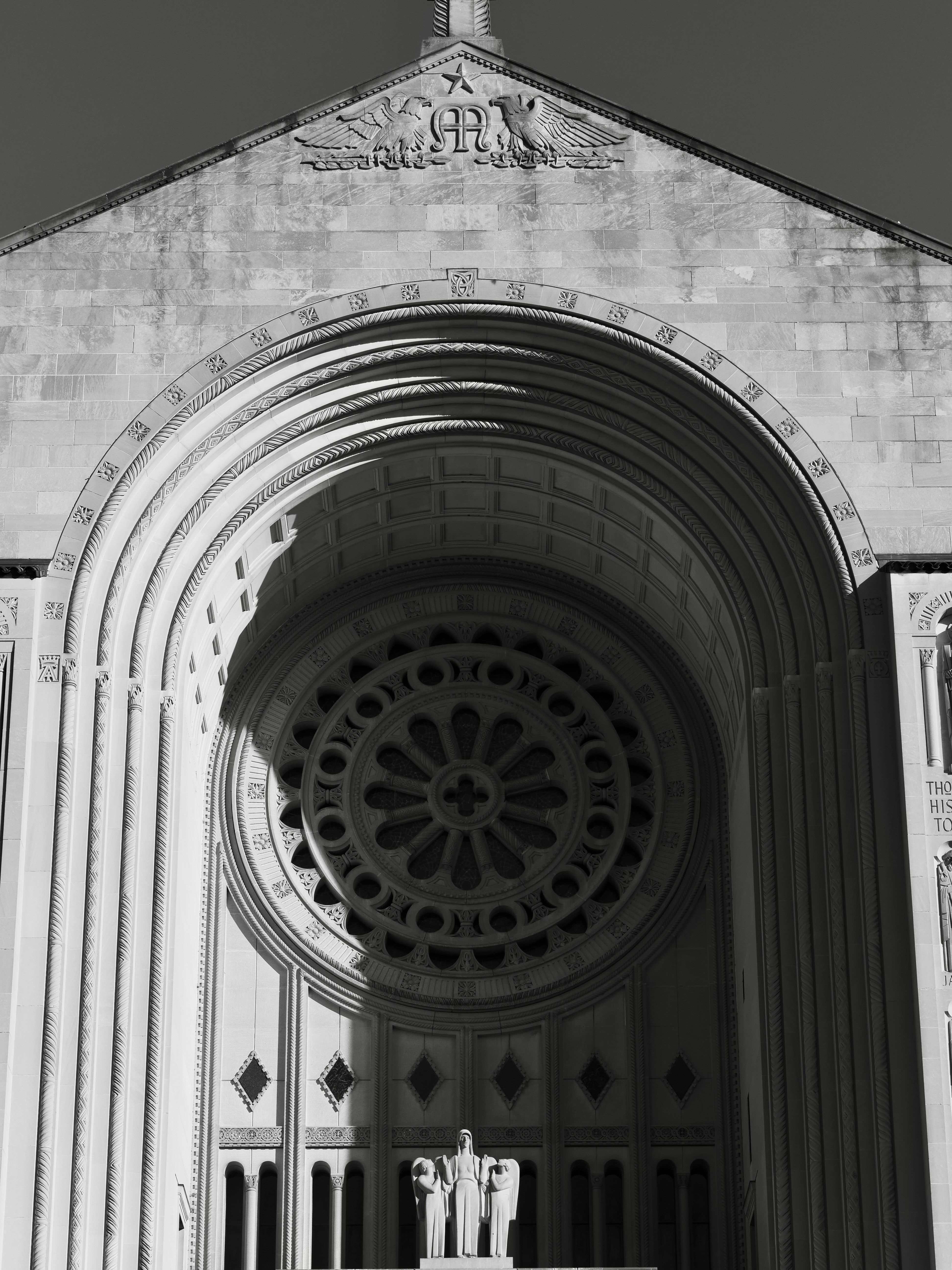 Monochrome facade featuring a monumental arched doorway with a circular rose window, flanked by sculpted figures, captured with emphasis on symmetry and architectural texture.