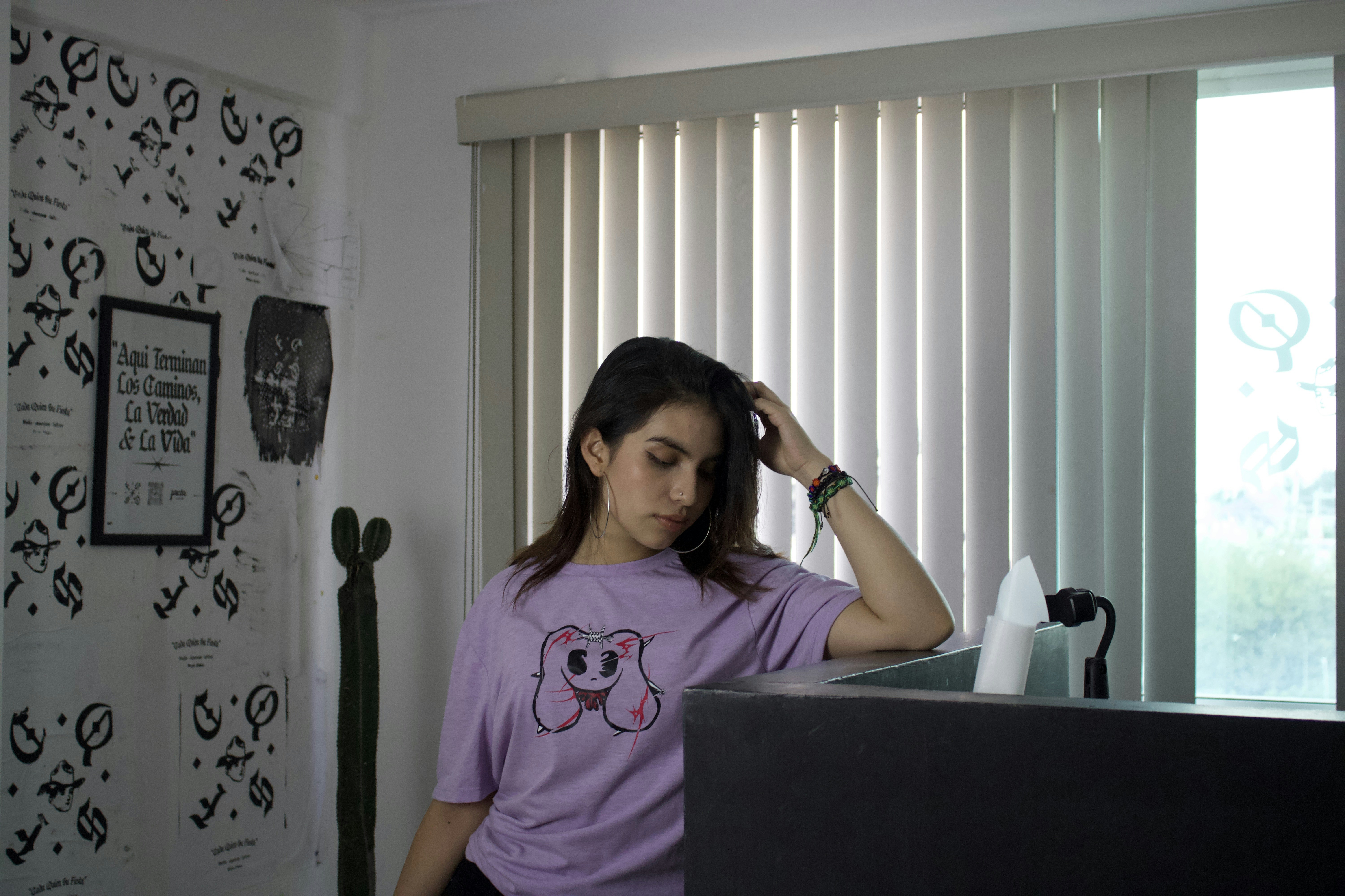Young woman in a lavender t-shirt rests her head on her hand beside a black counter in a softly lit room with patterned wall art and vertical blinds. A small, subtle backstage moment in a casual indoor setting.