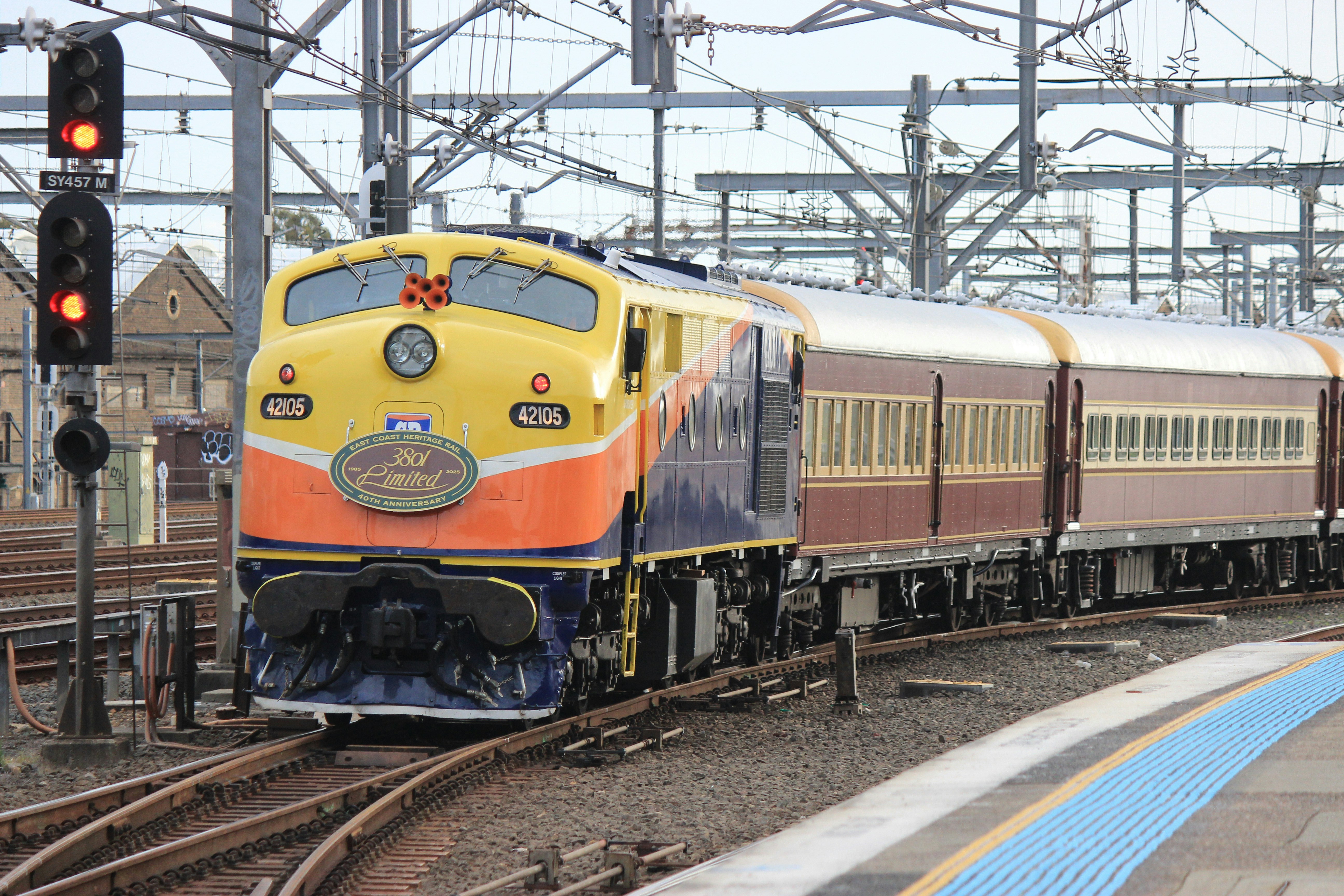 Bright diesel locomotive leads a multicolor passenger train past a station platform with blue tactile paving and overhead wiring.