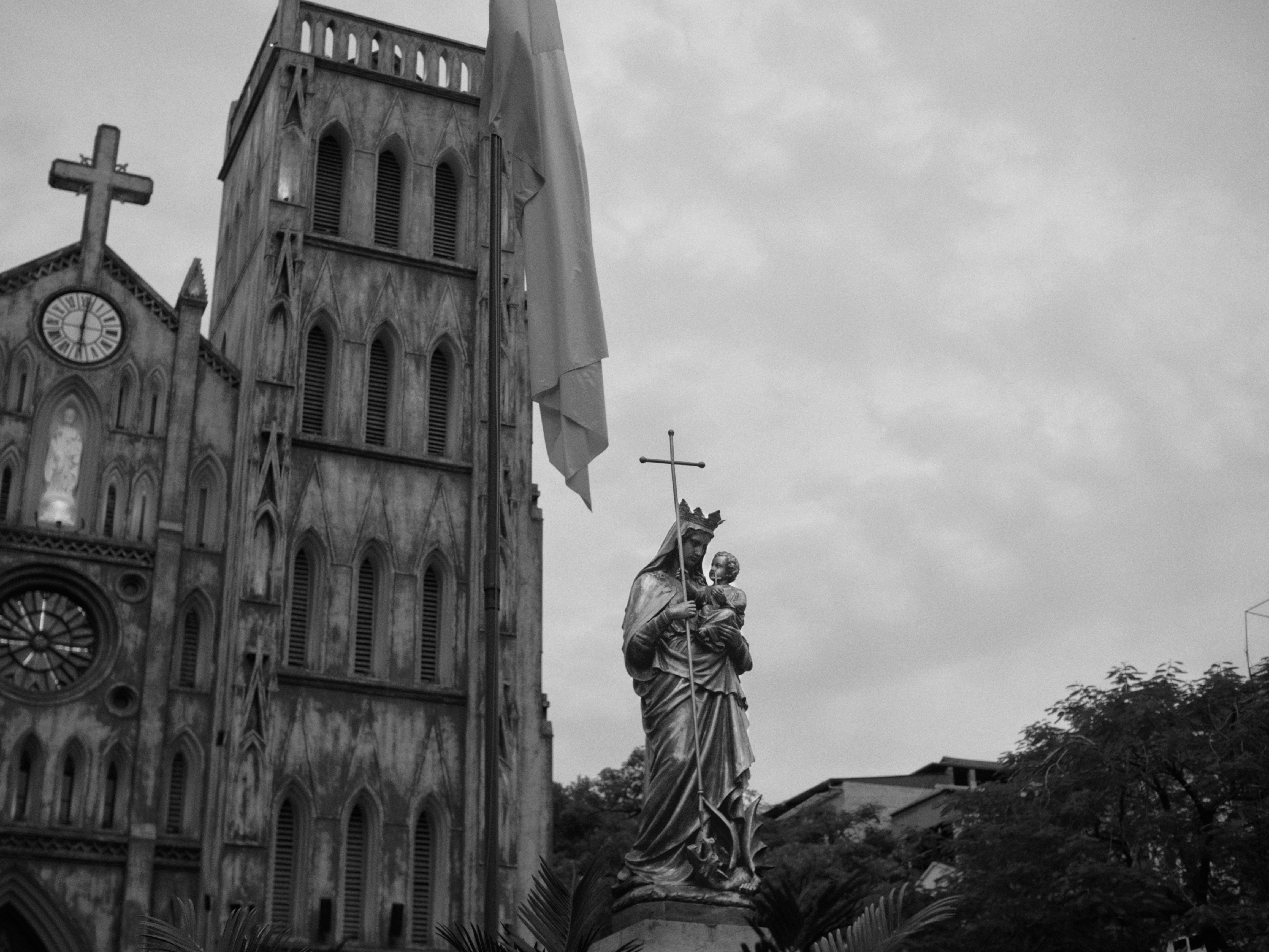 Grey-toned cathedral façade with a towering statue of a saint holding a child and a cross, flanked by arched windows and a clock tower; solemn, architectural scene.