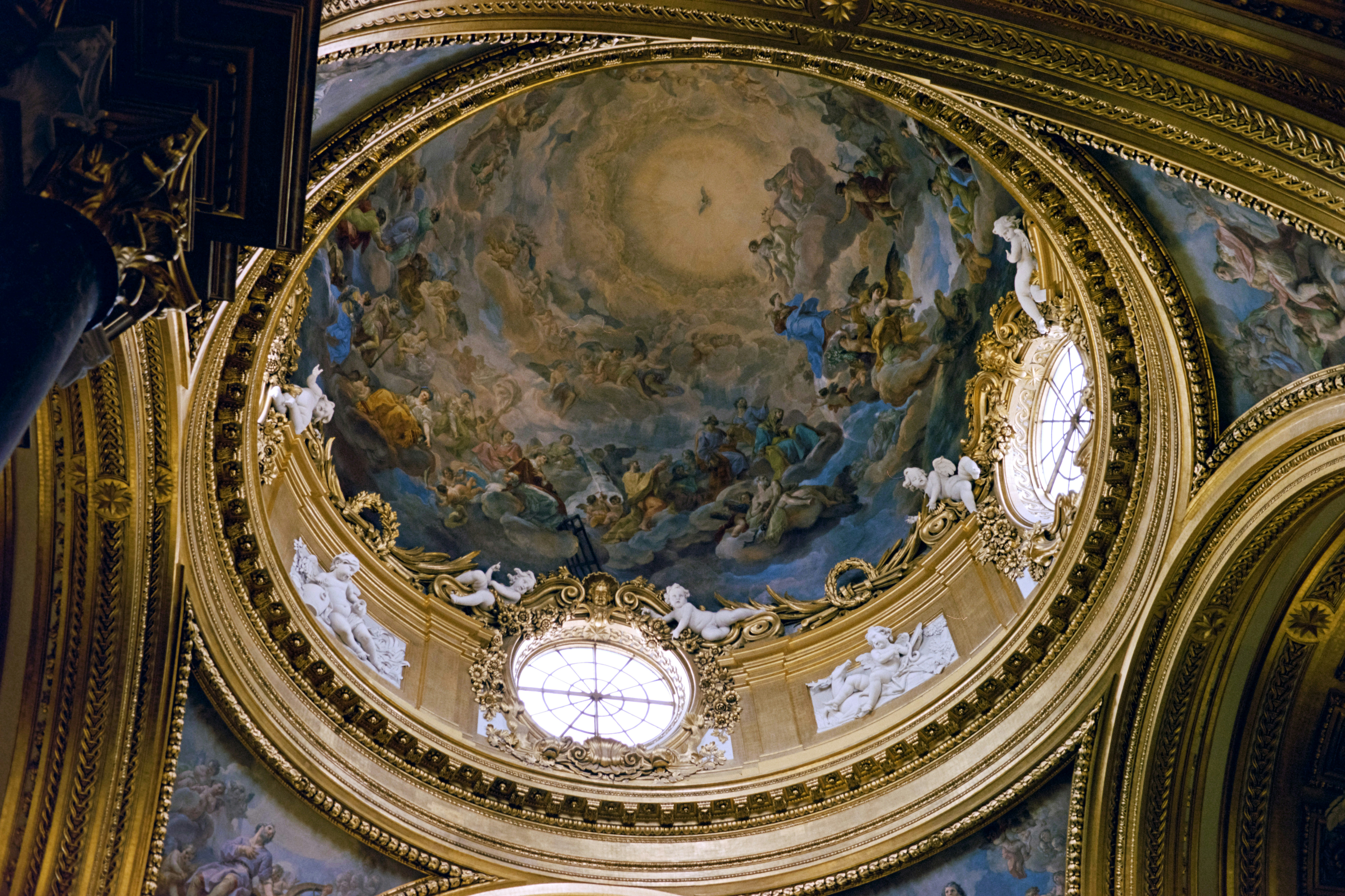Ornate gilded dome interior with painted celestial scene and circular windows, highlighting baroque architecture and sculpture.