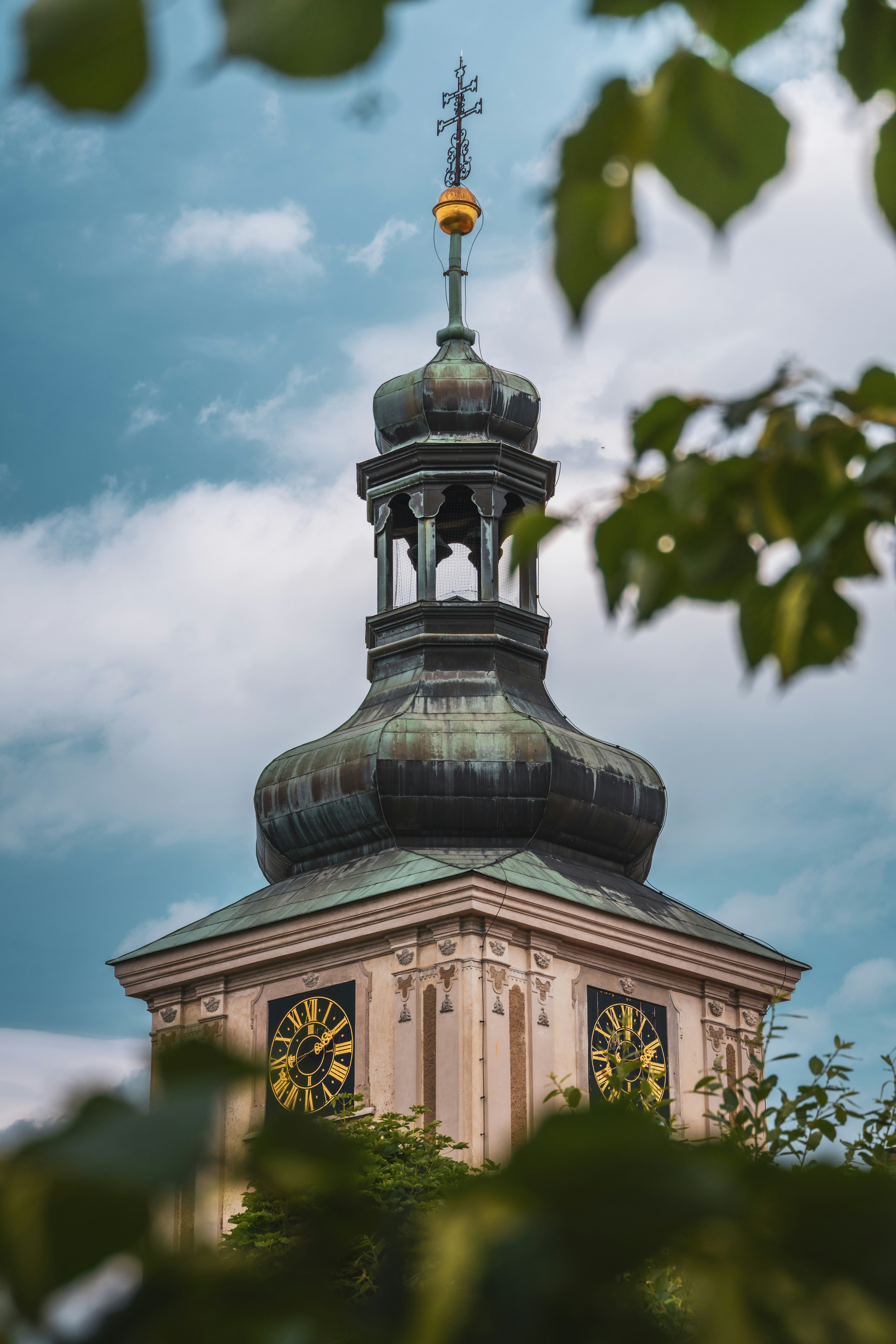 Stone church tower with a green patinated copper dome, ornate clock faces, framed by foliage against a bright blue sky.