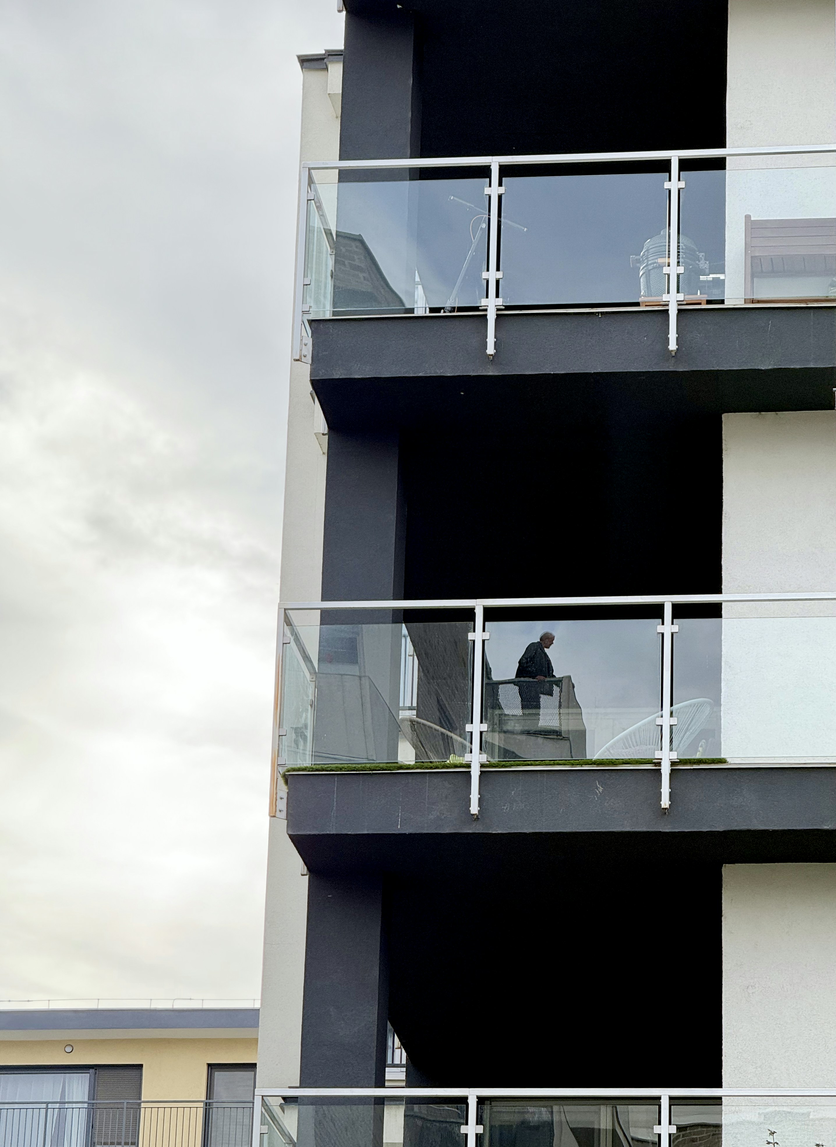 A person sits at a balcony with glass railings on a modern apartment building, framed by vertical concrete and dark recesses; the scene captures a quiet urban moment. This is a photograph of architectural space.