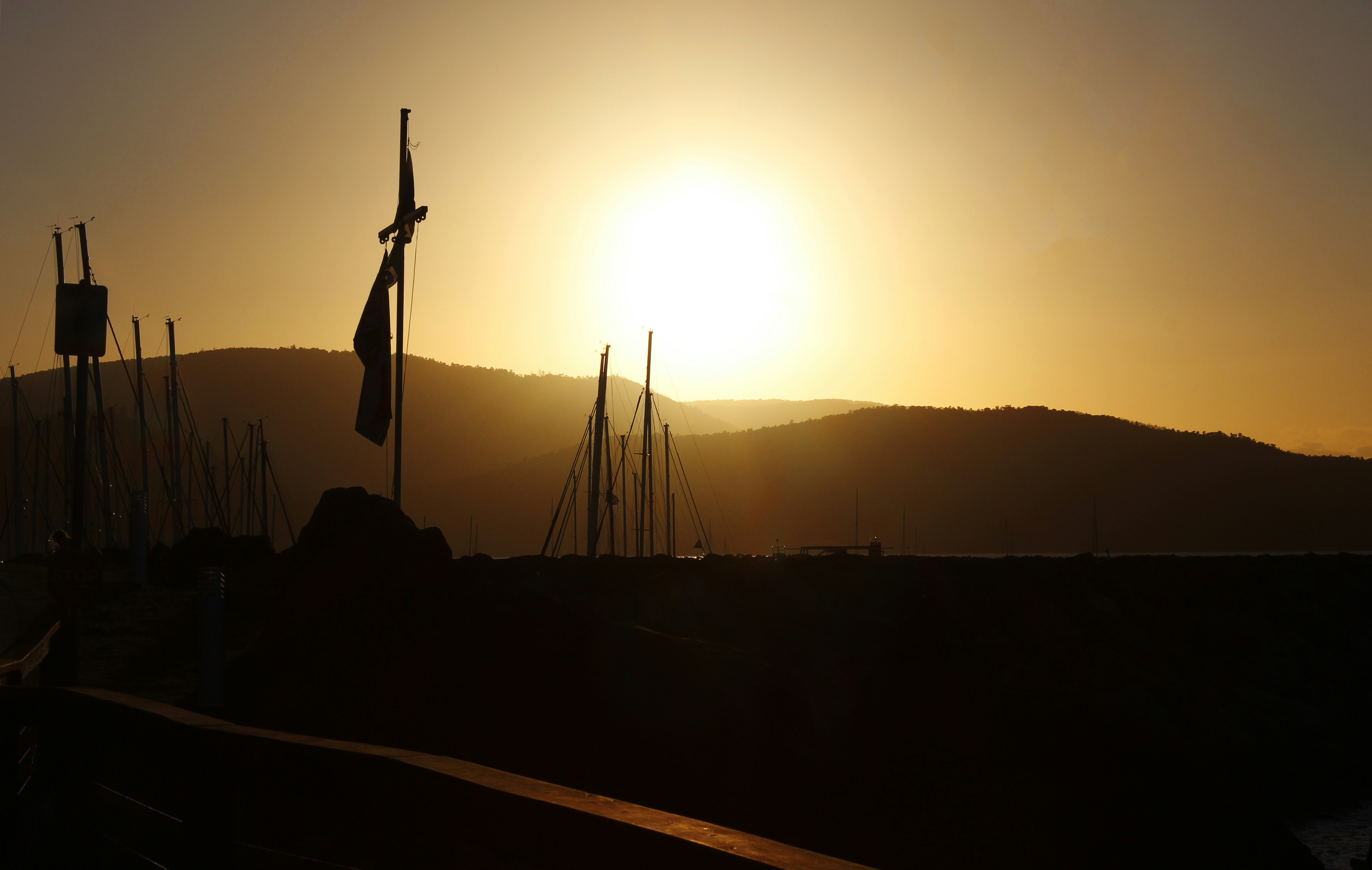 Sunset over a harbor with silhouettes of boats and masts along a dark shoreline.