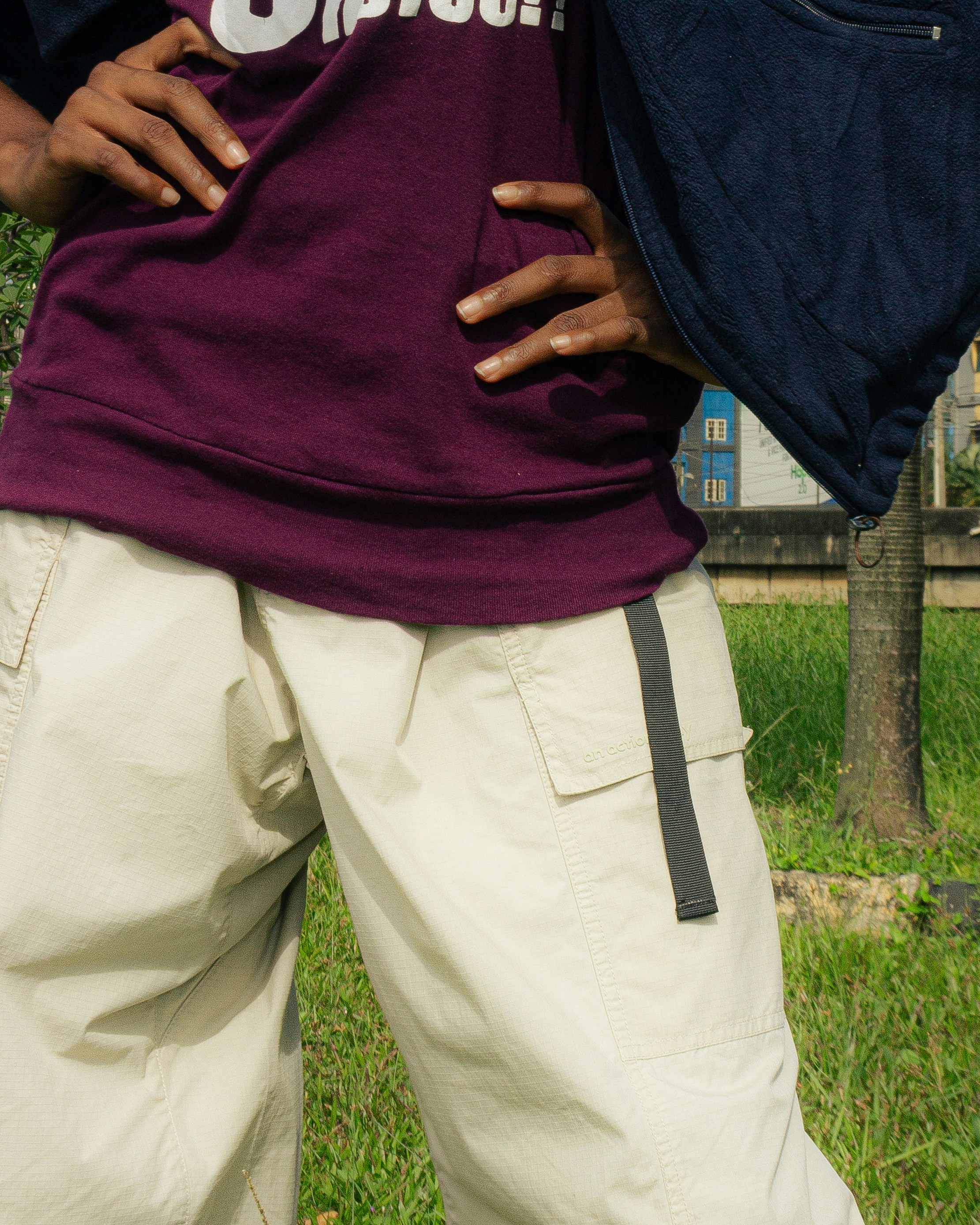 Close crop of a person wearing a maroon shirt and beige cargo pants with a dark strap hanging from a pocket, standing on grass near trees and a bench.