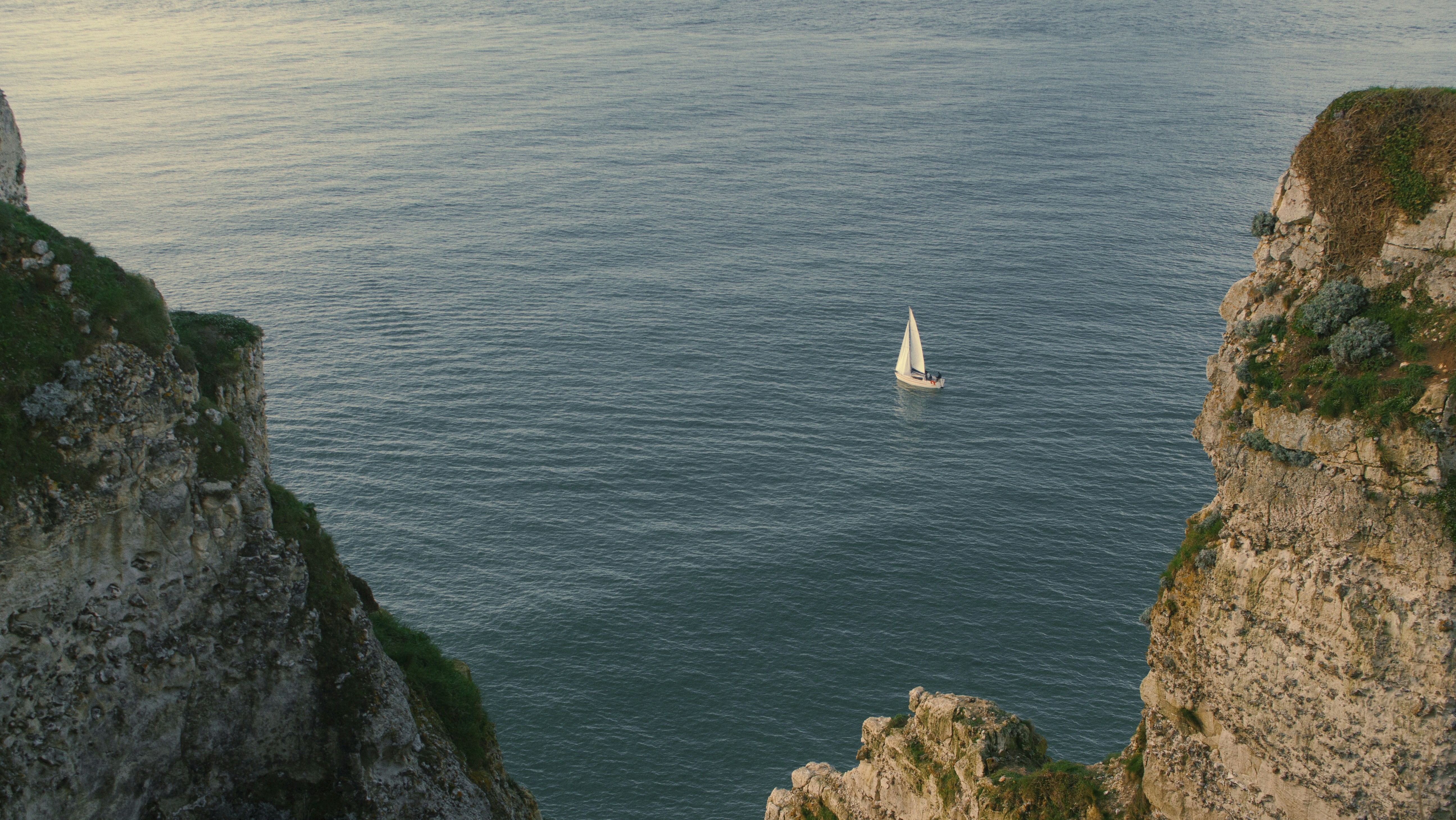 A lone sailboat glides between towering coastal cliffs over calm blue water, with rugged rock faces framing the scene.
