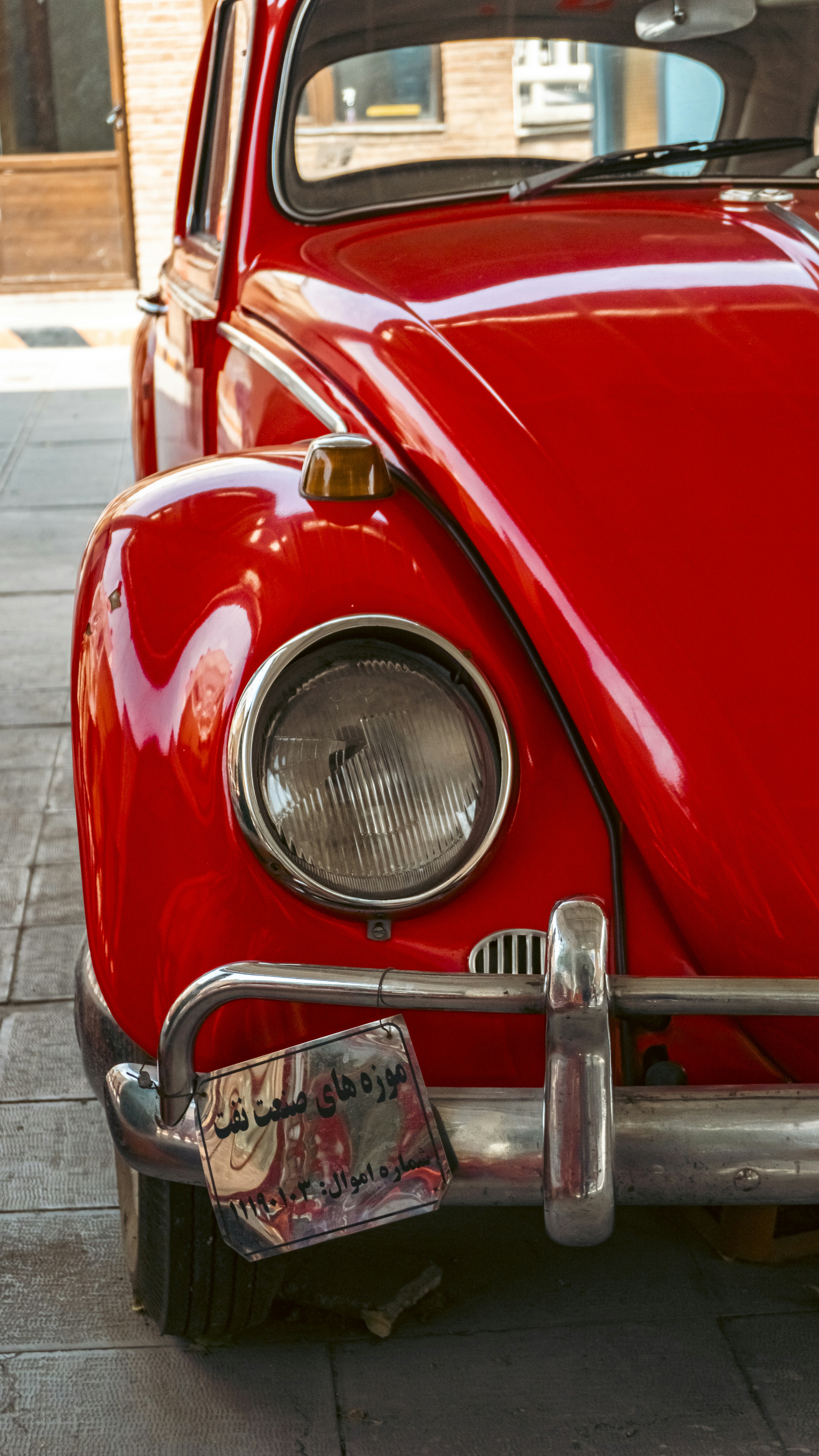 Close-up of a glossy red vintage car front with round headlamp, chrome bumper and a tilted license plate, capturing the craftsmanship and reflections of an urban street scene.