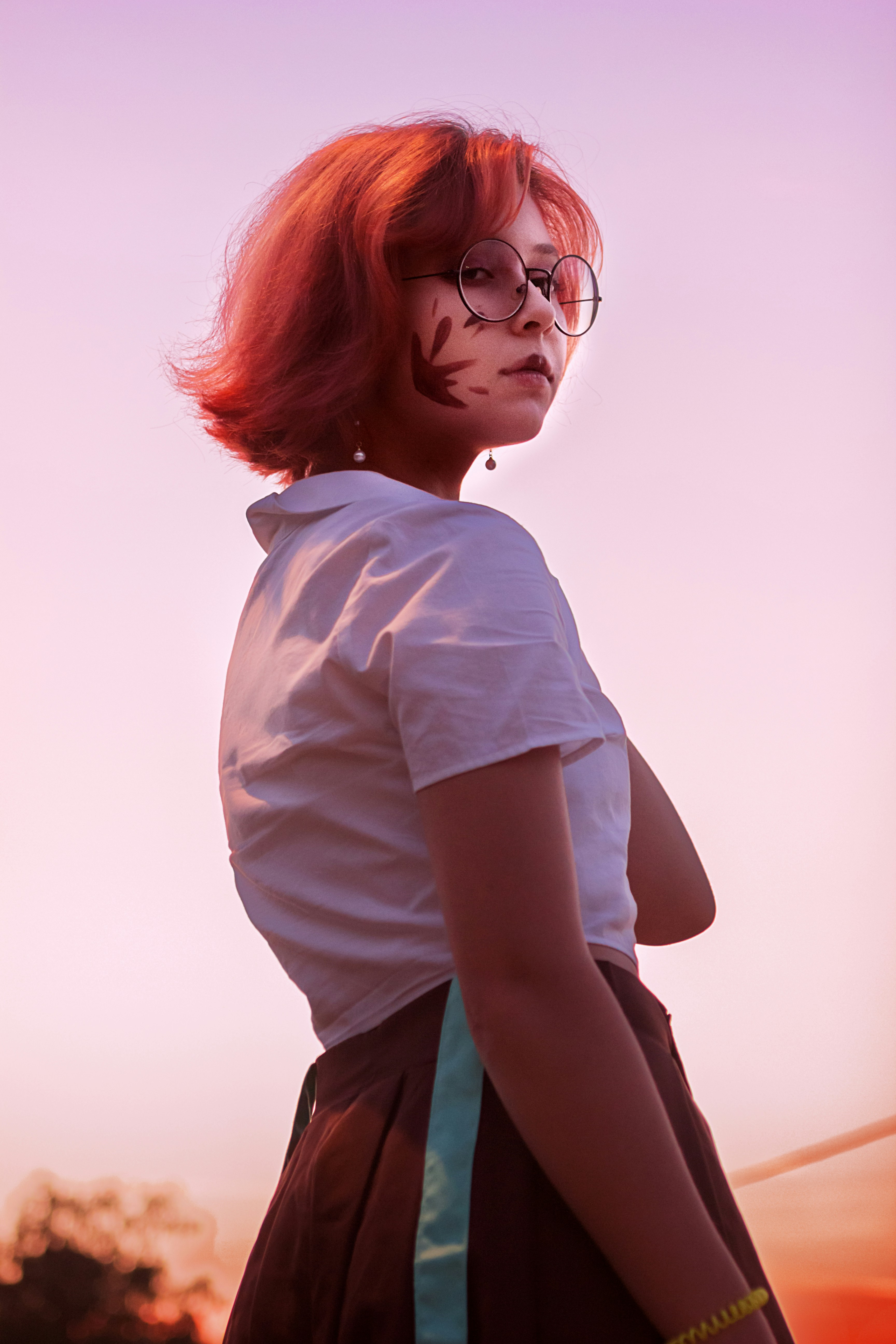 A young woman with red hair and round glasses gazes upward, her face marked with a decorative pattern, as the sunset casts a warm, pinkish glow over her white blouse and teal ribboned skirt.