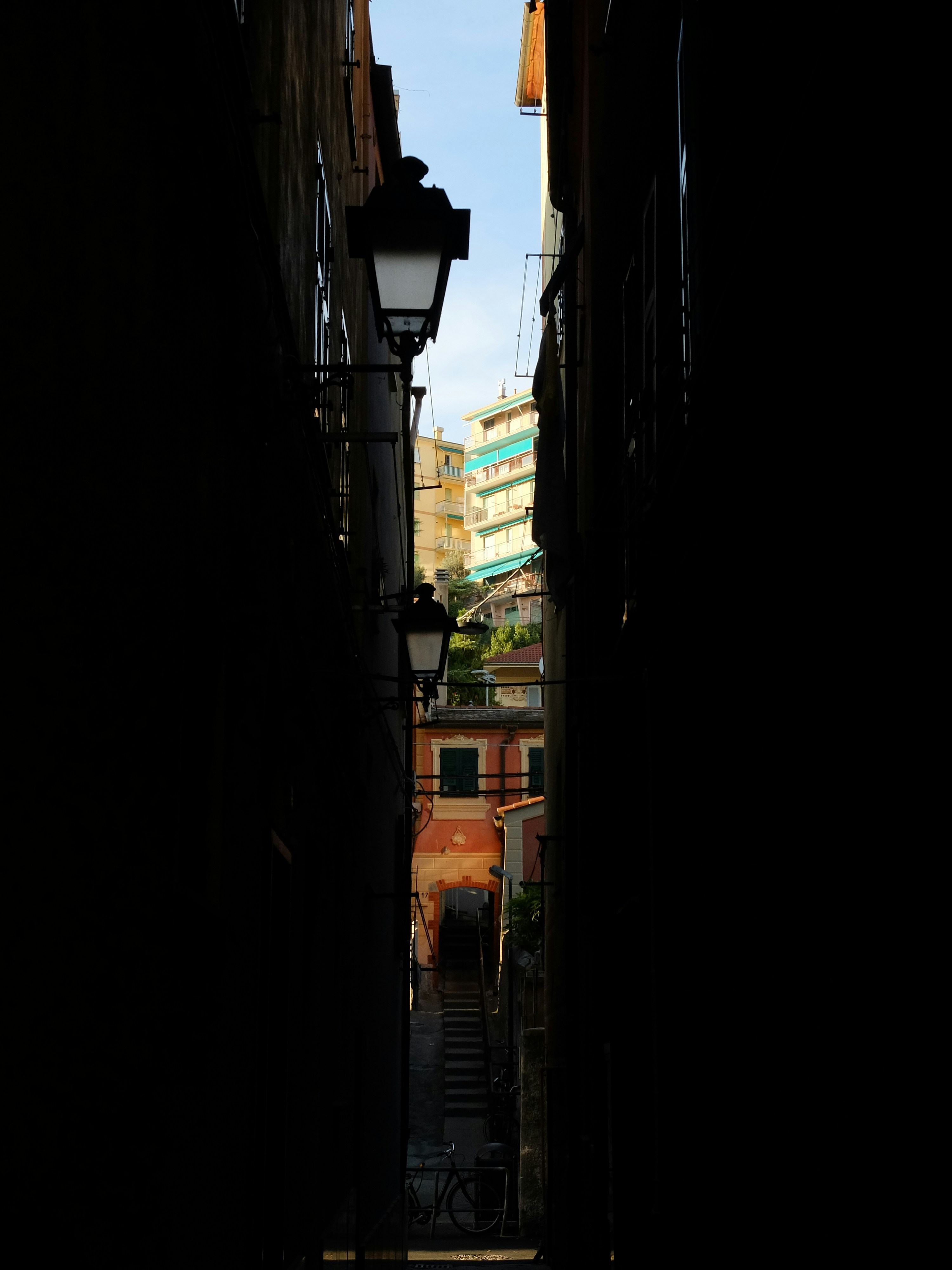 A sunlit courtyard rises beyond a dark, narrow alley framed by tall buildings and classic street lamps.