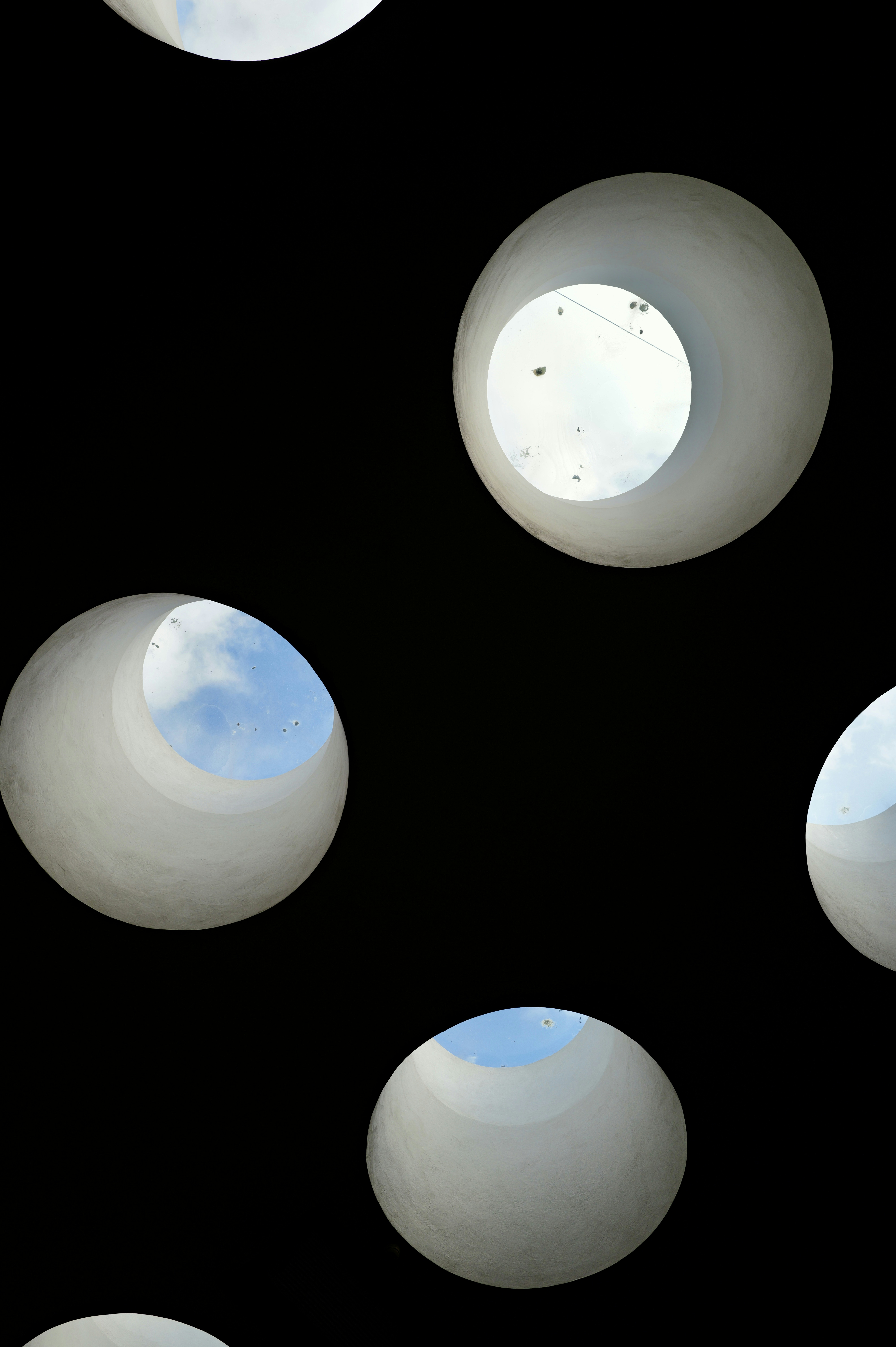 White cylindrical voids puncture a dark backdrop, revealing glimpses of blue sky and scattered debris. A minimalist architectural composition exploring negative space and geometric repetition.