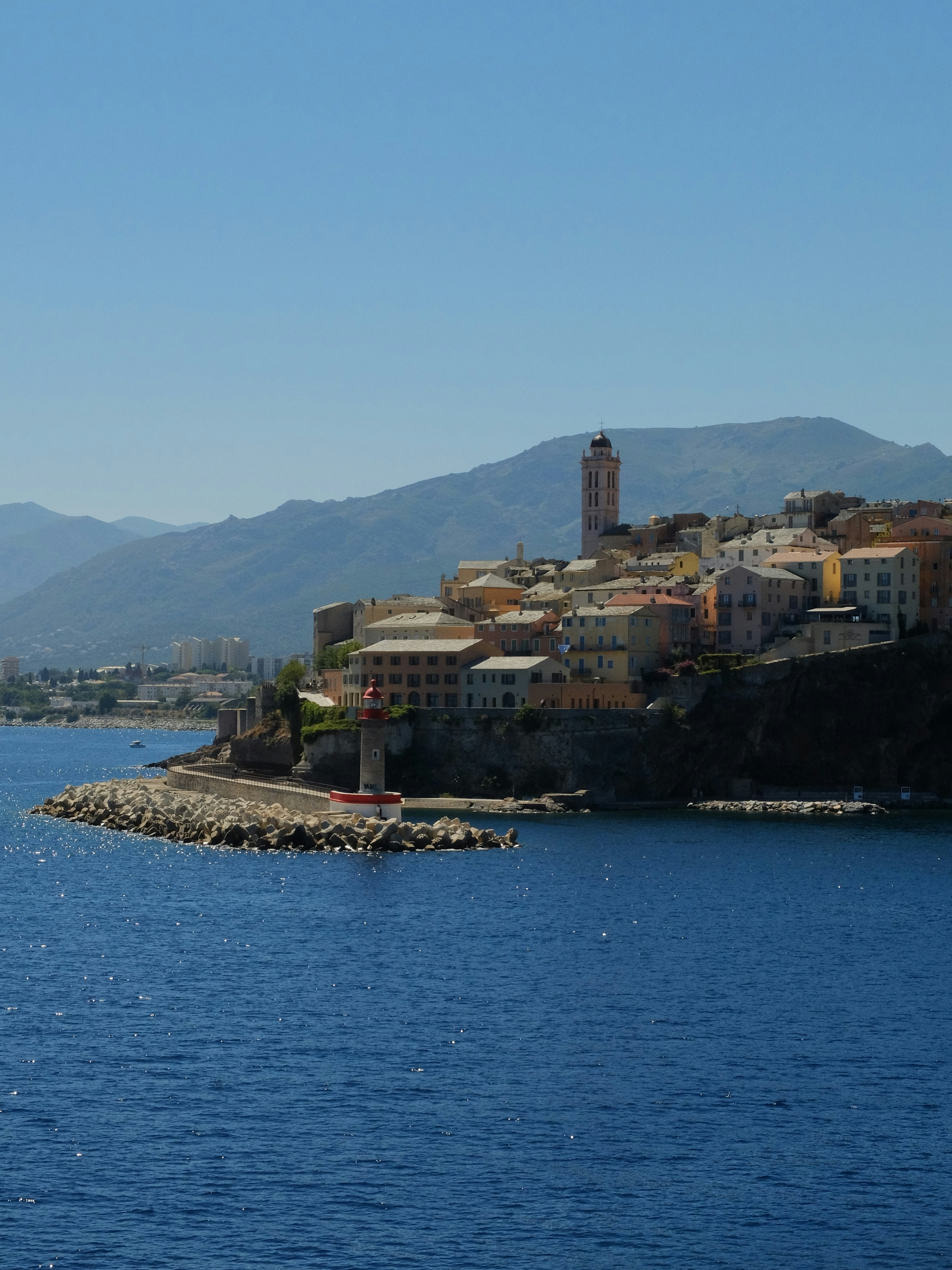 Sunlit coastal village perched on a rocky promontory with a tall tower overlooking the harbor, framed by distant mountains and bright blue sea.