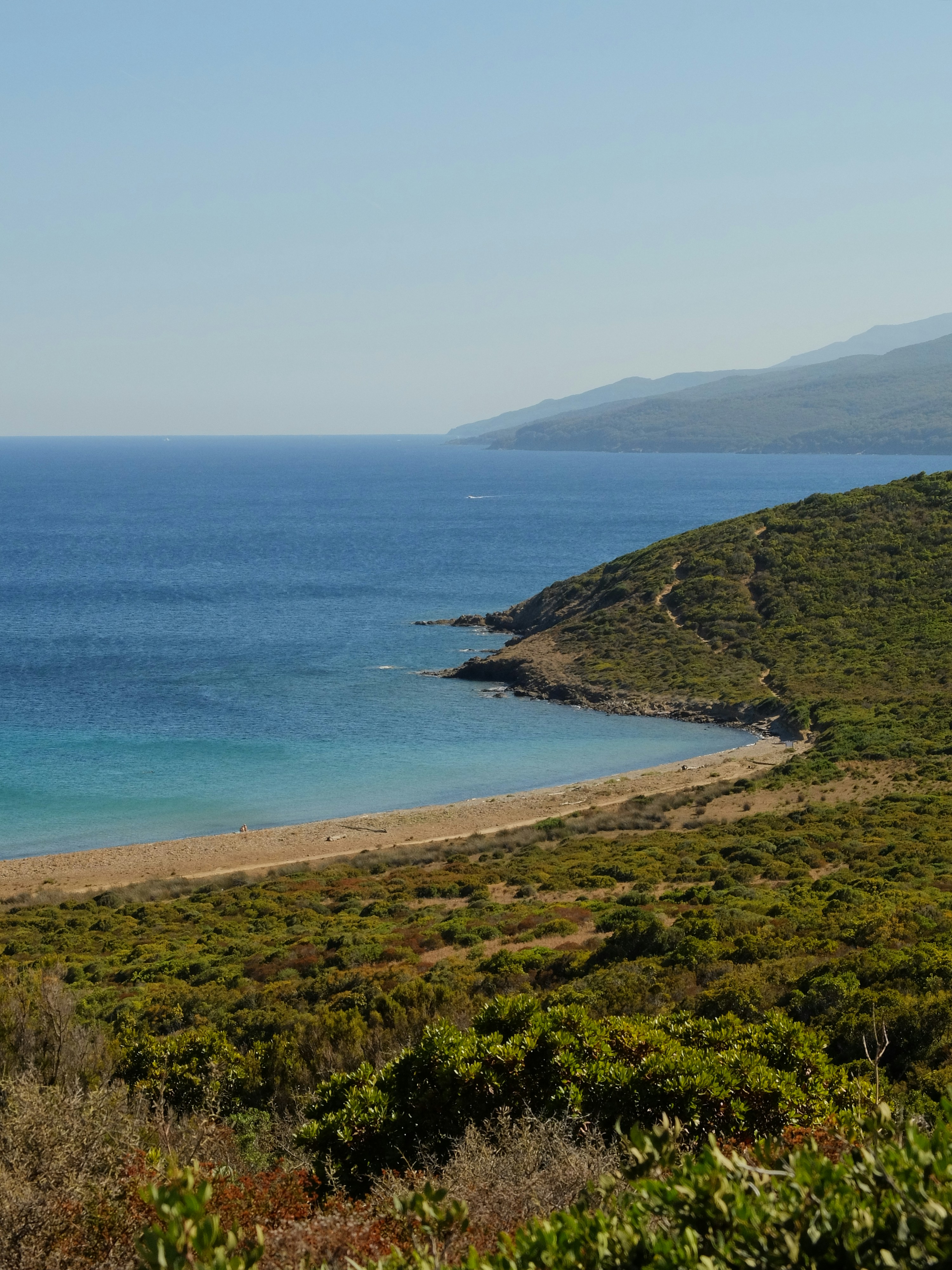 Coastal landscape with turquoise bay and rugged hillside meeting a sandy beach; distant headlands fade into blue haze, foreground shrubs adding texture. A serene sea-side scene emphasizing natural shoreline and terrain.