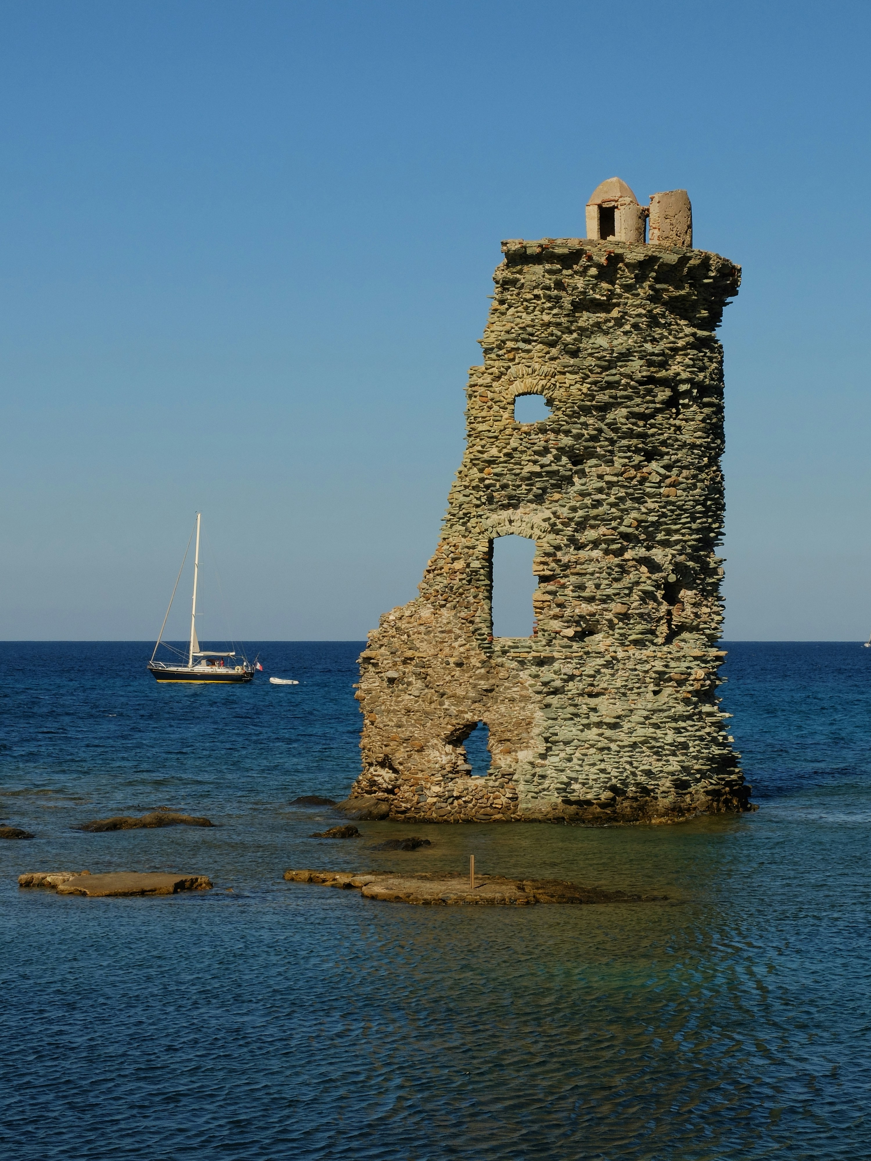 Rugged stone tower stands in the shallow turquoise waters as a lone sailboat glides near the horizon, under a clear blue sky.