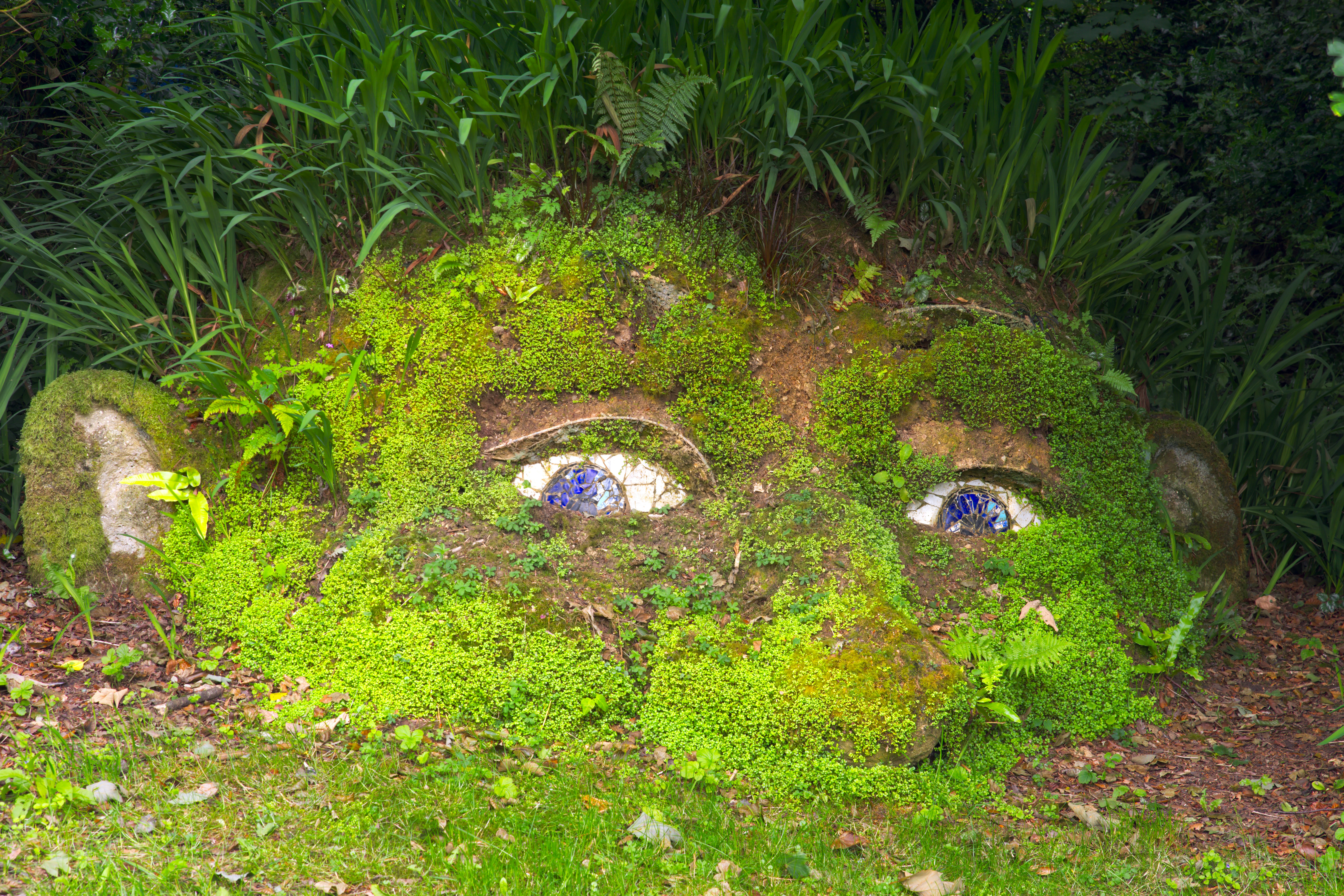 Mossy stone sculpture emerging from soil with bright blue eyes peering through greenery; a nature-made face framing the scene.