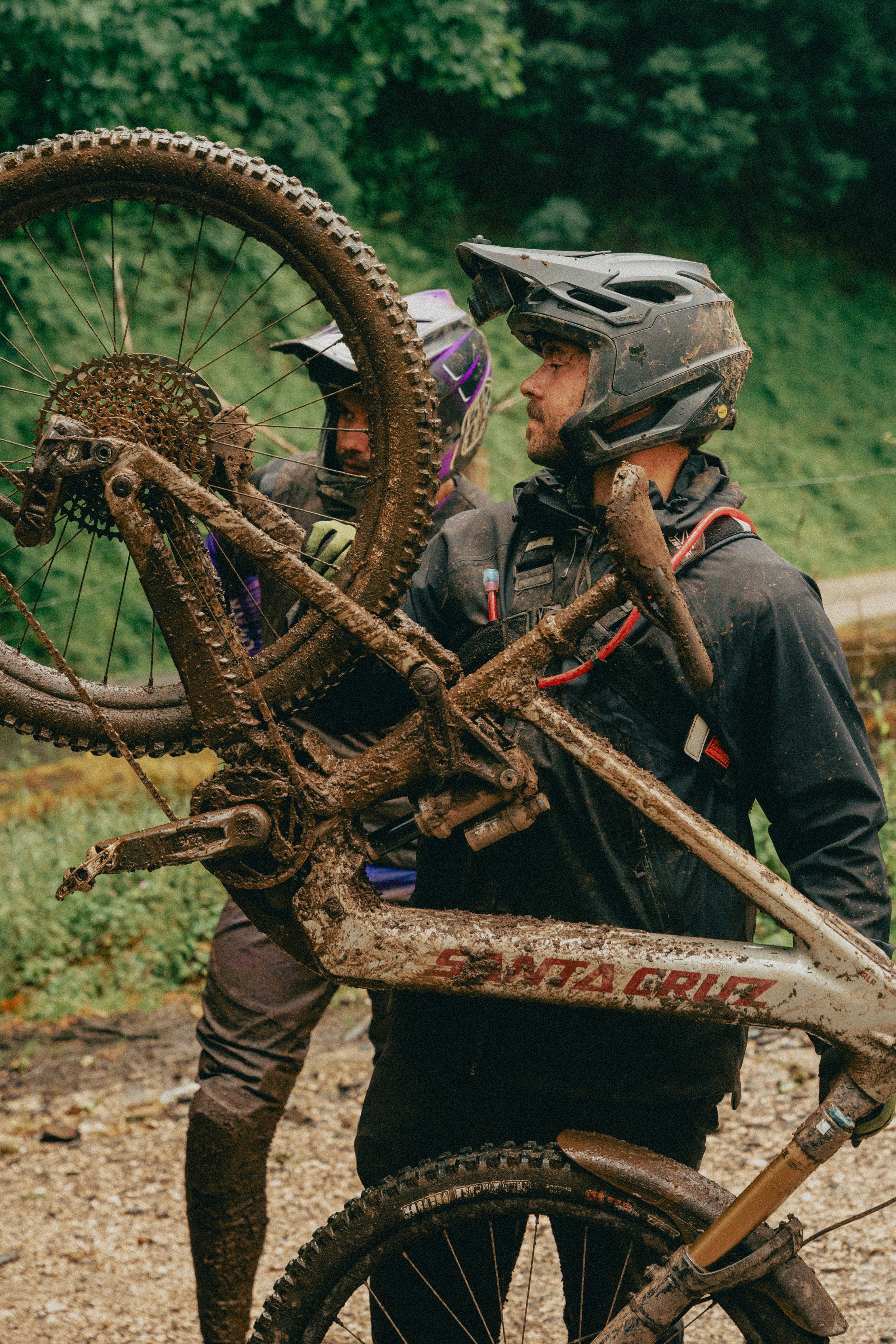 Mud-Splattered Recovery: Two Riders Lift a Stalled Mountain Bike in a Forest Trail