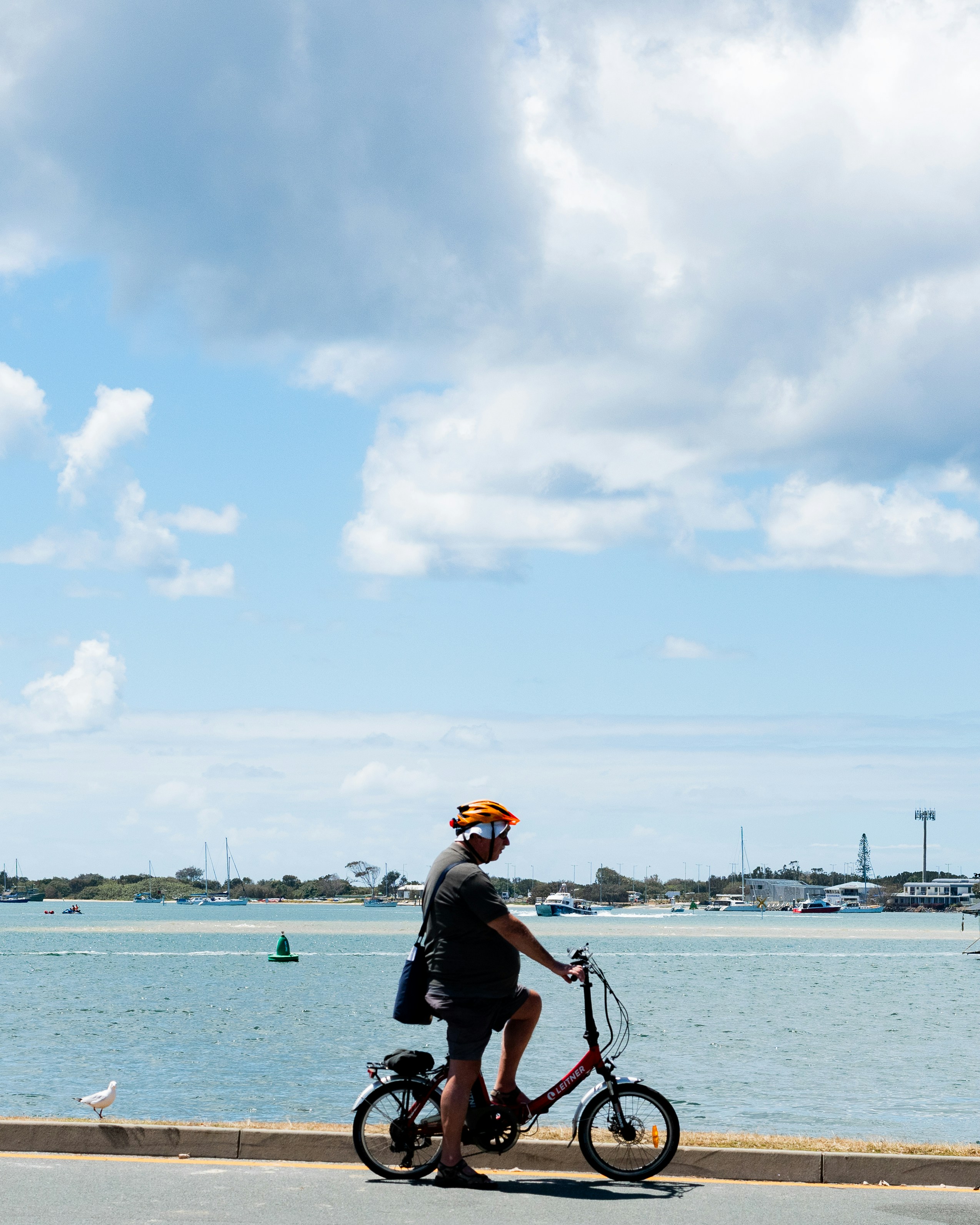 A man wearing a helmet rides a folding bike along a seawall with calm water and boats in the distance under a bright sky.