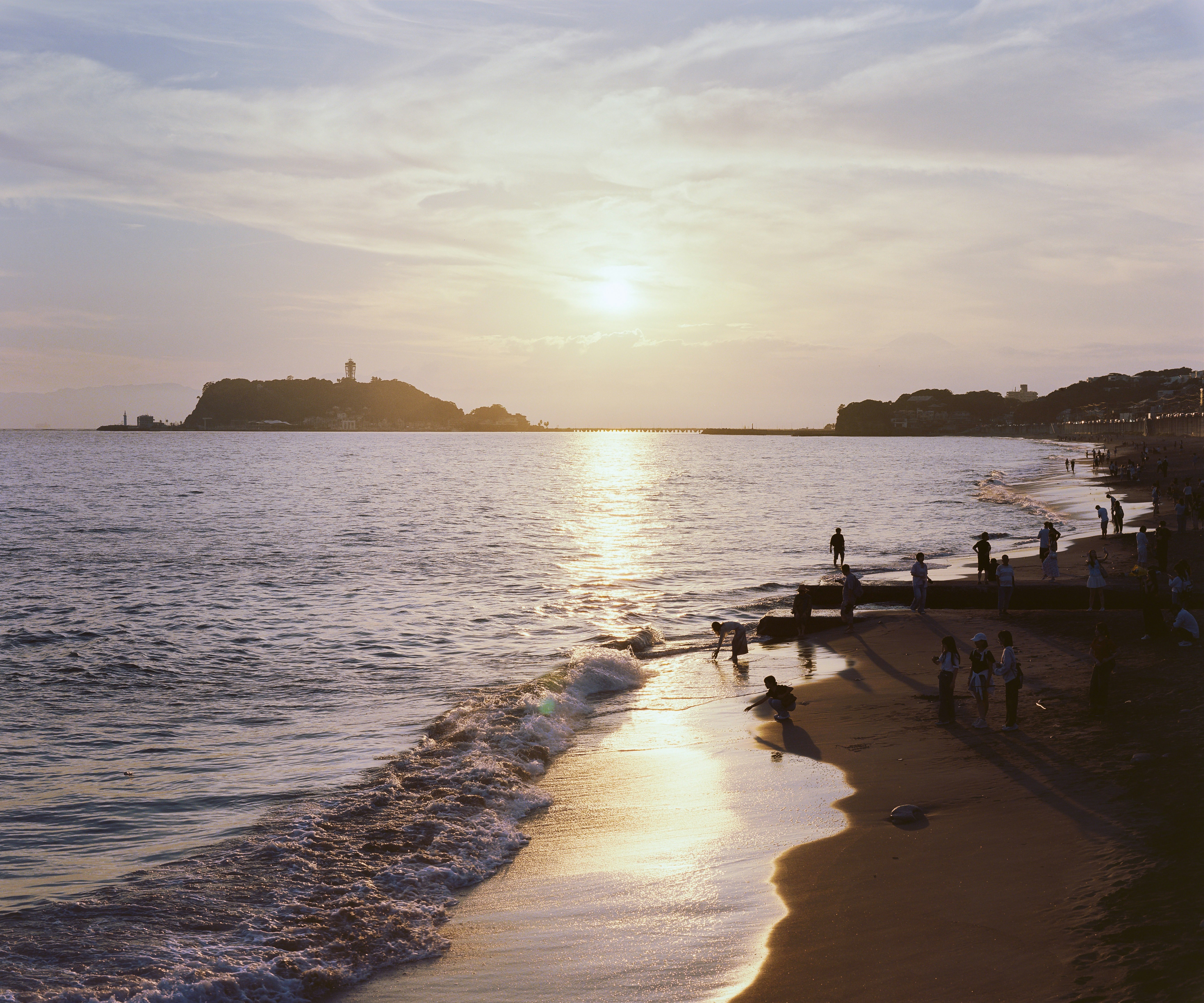Sunlit coastline with gentle waves and a distant lighthouse on an island; people along the shore and jetty during golden hour. A serene seaside scene emphasizing calm light and activity at the water's edge.