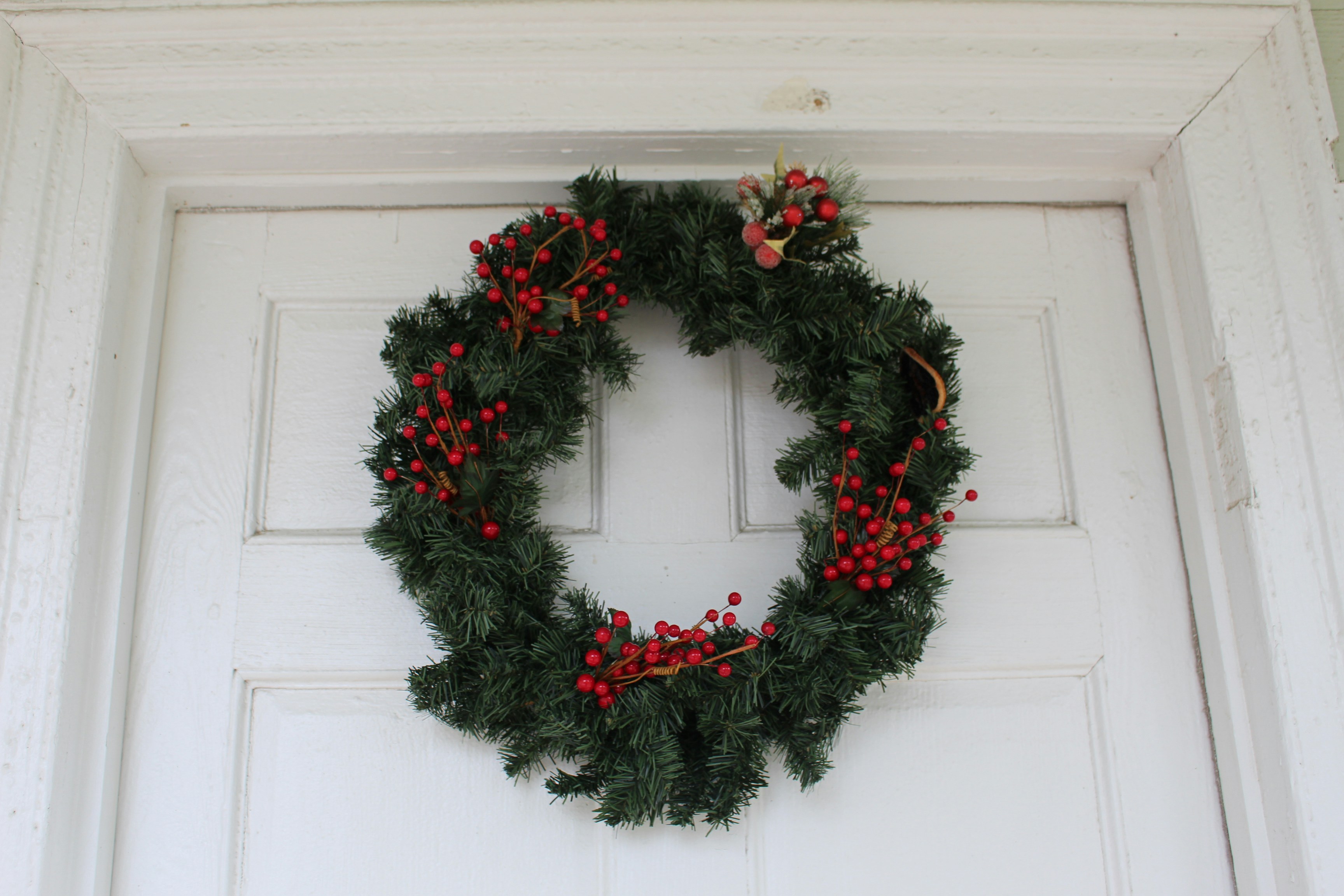 A festive evergreen wreath with red berries hangs on a white paneled door, centered to convey a simple holiday welcome.